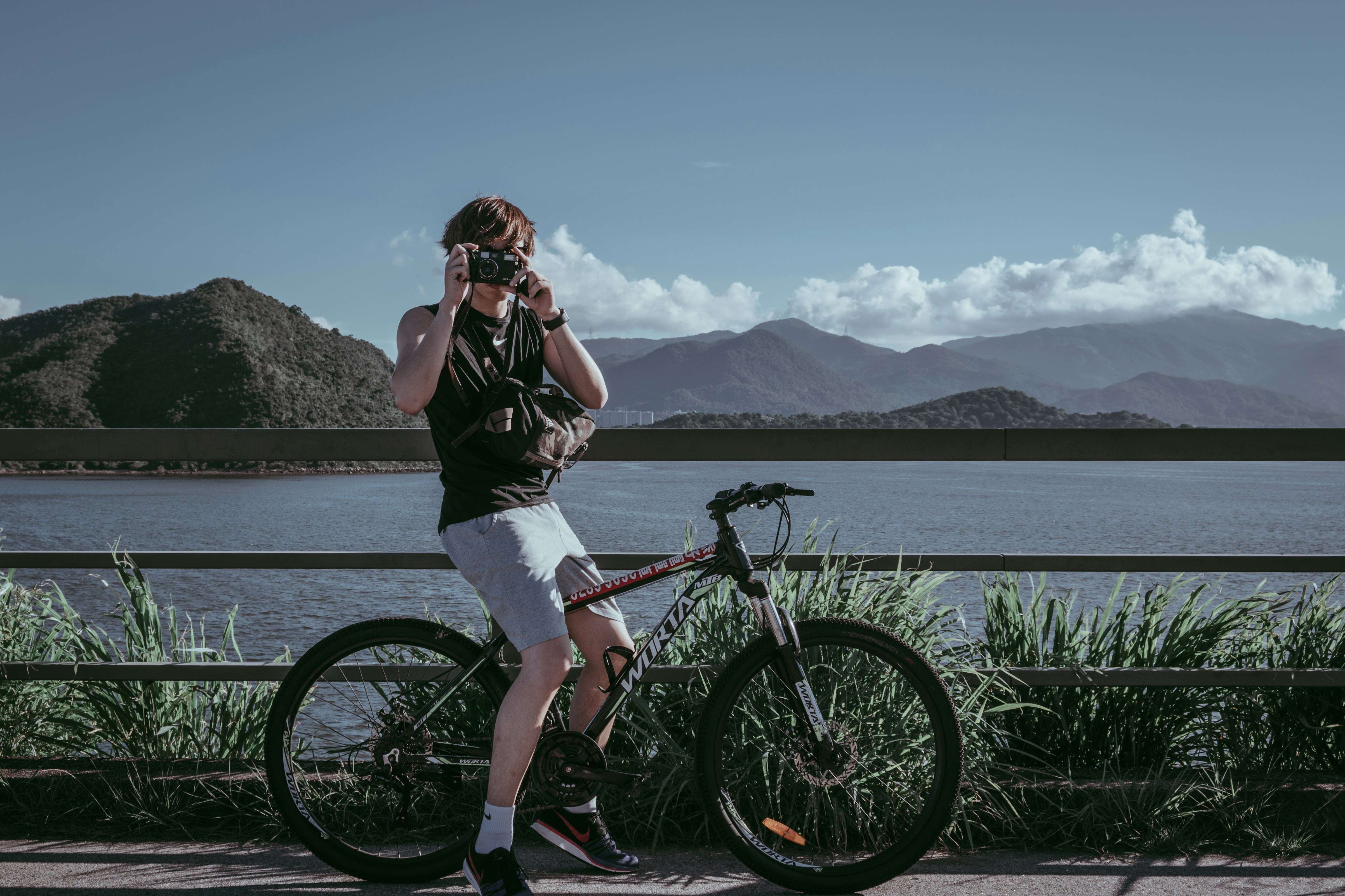 A cyclist pauses by a serene lake, capturing the landscape with a camera, framed by mountains and lush greenery.