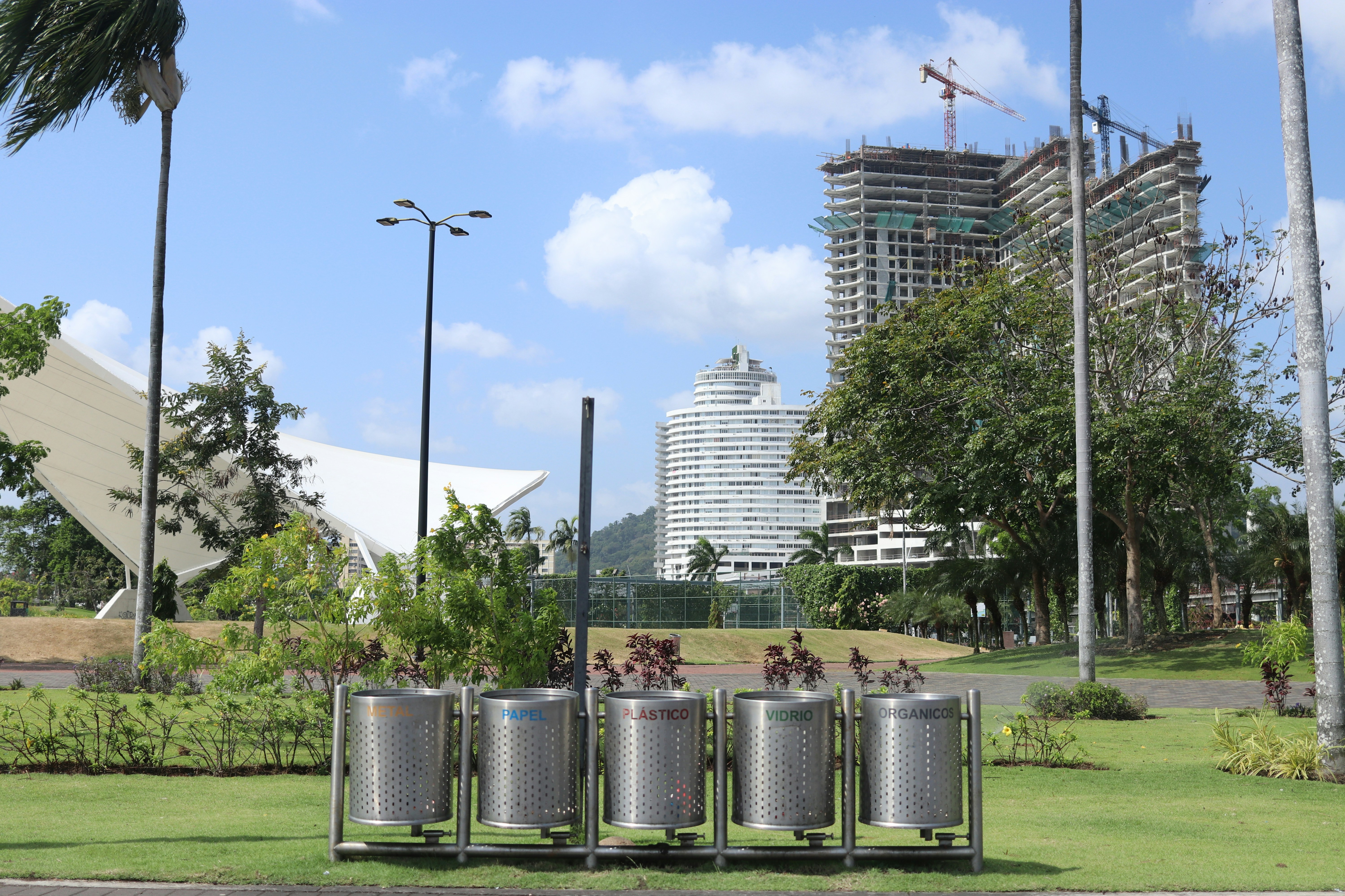 Five sleek trash bins positioned in a lush green park, framed by contemporary buildings and a clear blue sky.