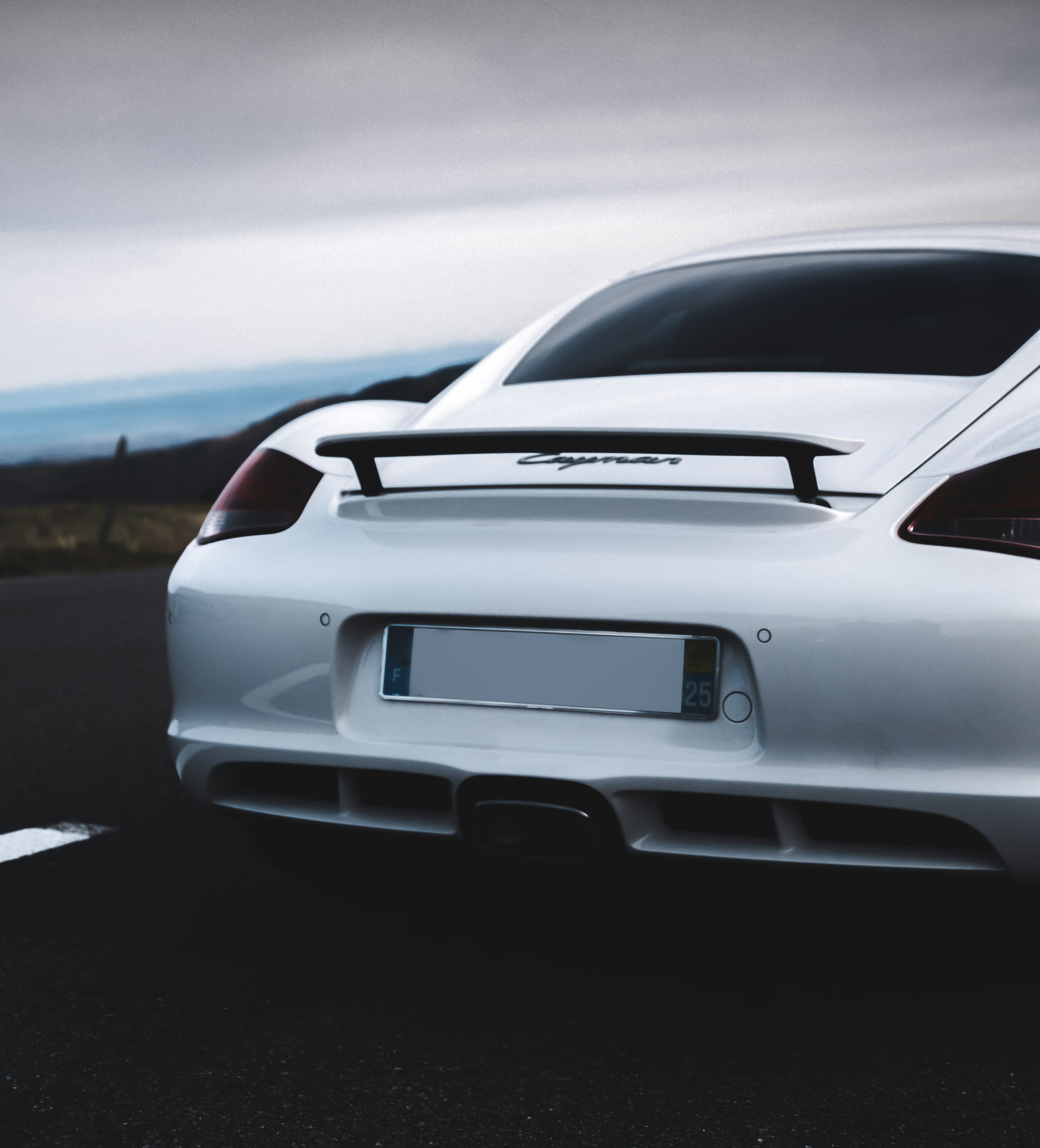 Sleek white Porsche Cayman parked on a winding road with a dramatic sky in the background.