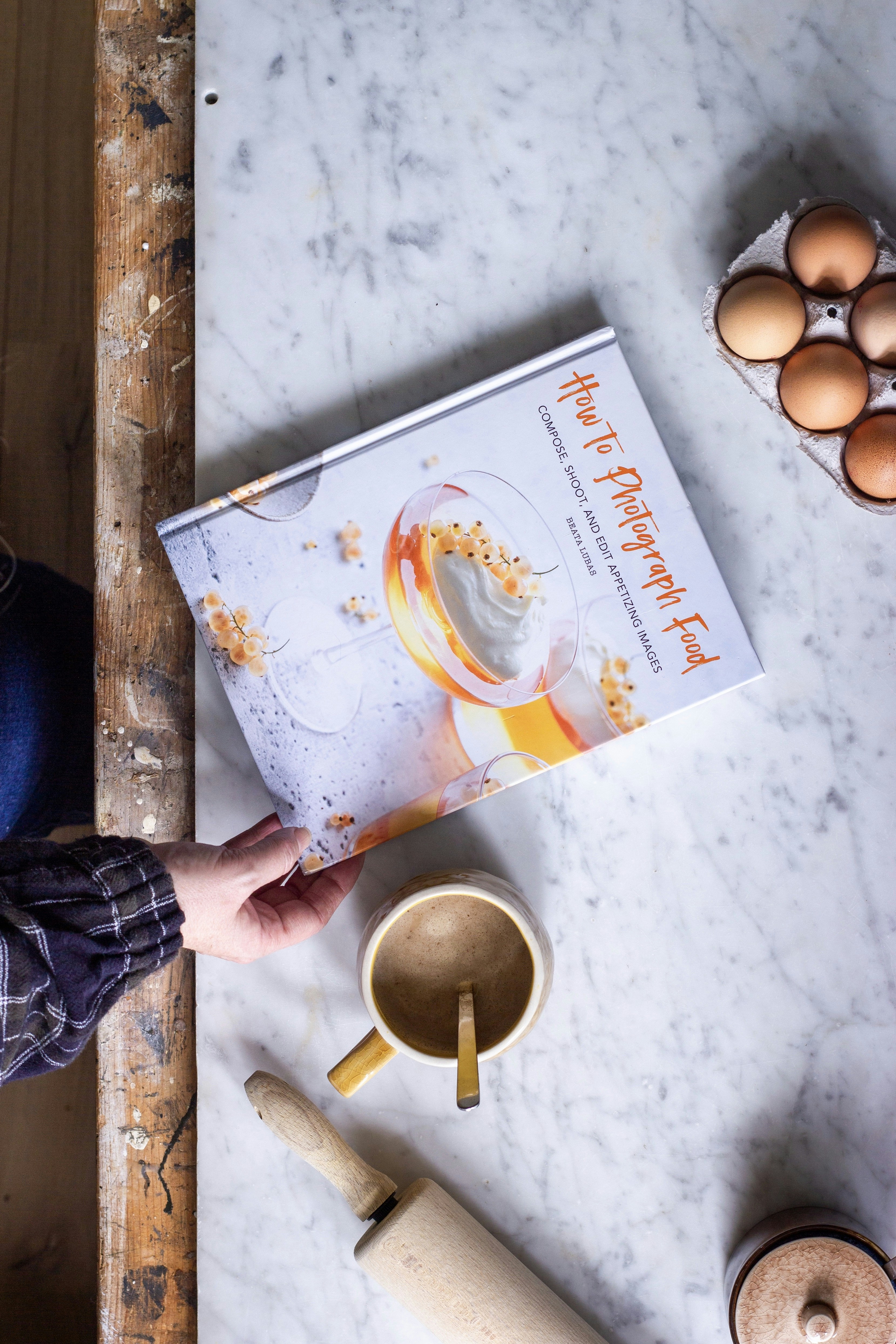 A hand reaches for a cookbook titled 'How to Photograph Food' resting on a marble table, accompanied by a bowl and eggs.