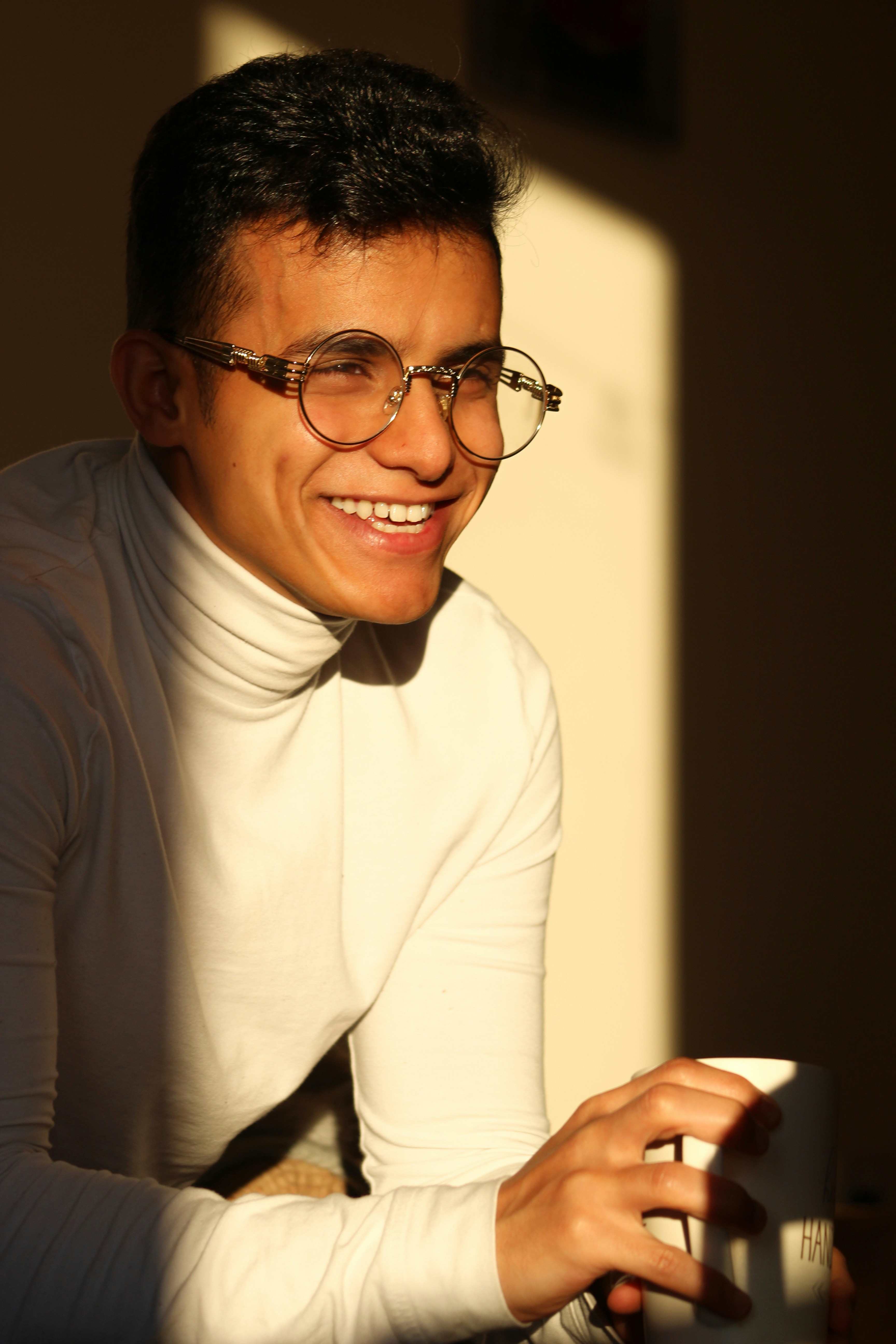 A young man in a turtleneck smiles warmly while holding a mug, illuminated by soft morning sunlight streaming through a window.
