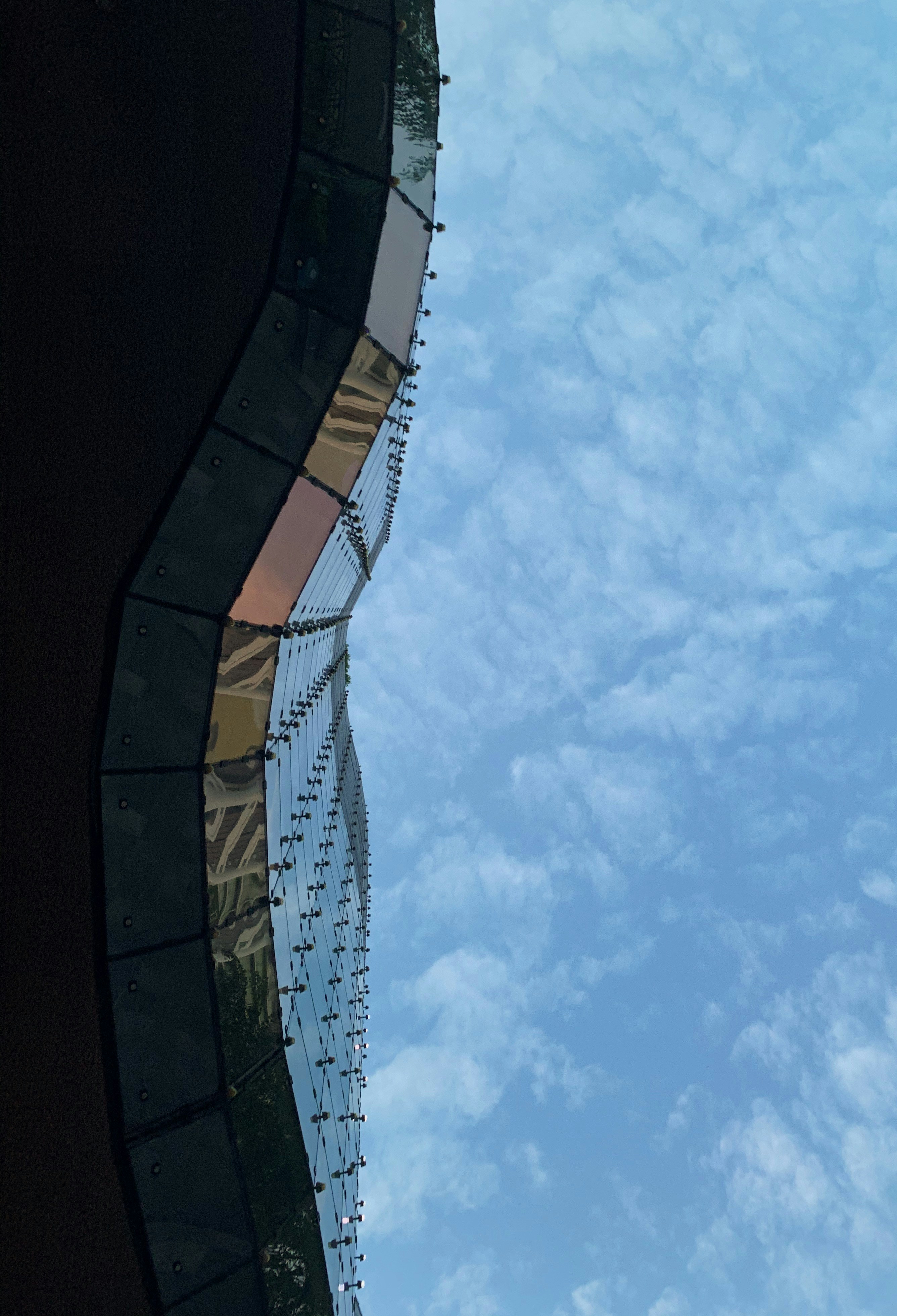 Curved glass facade reflecting the sky, framed by intricate metalwork and a backdrop of scattered clouds.
