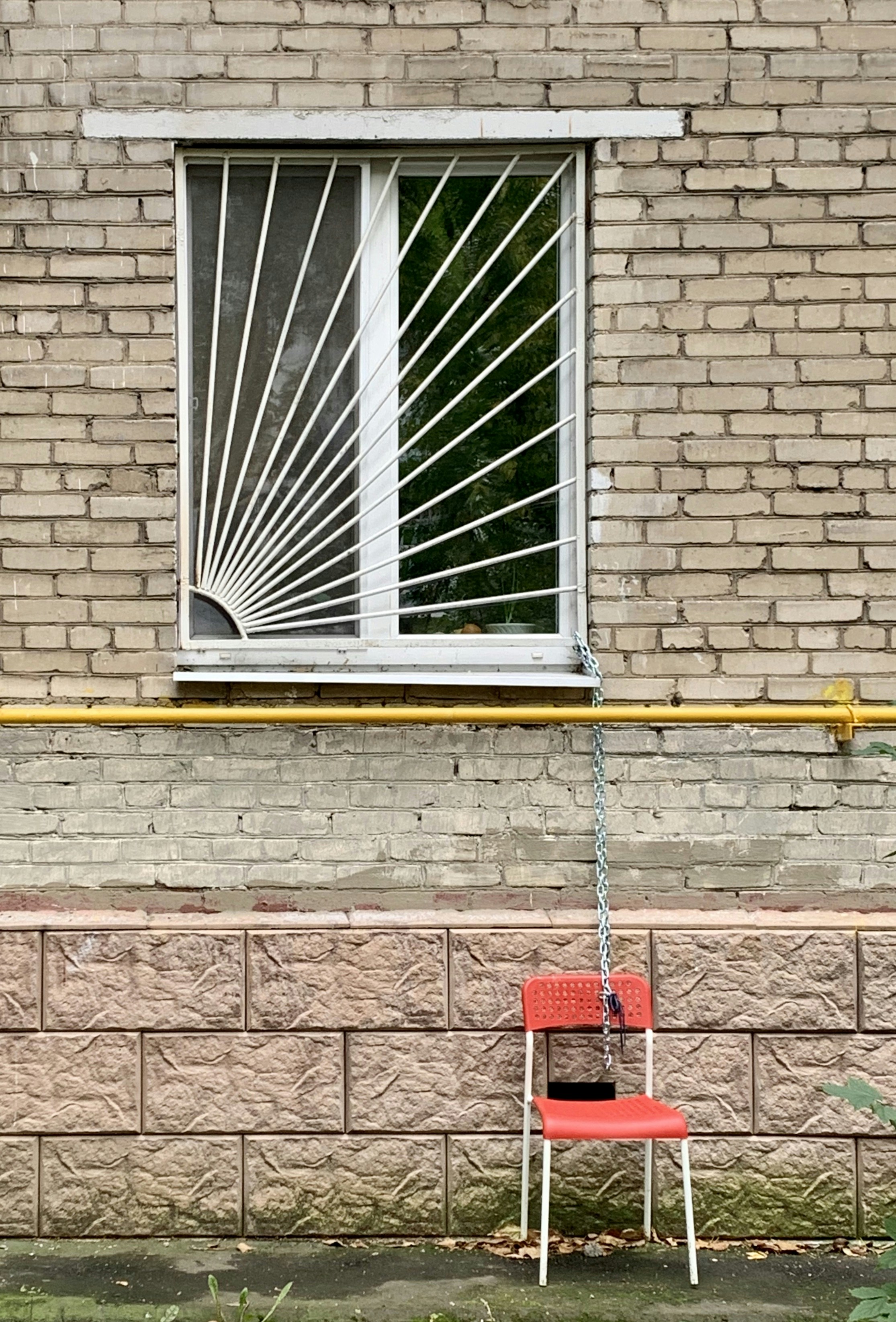 A chained red chair hangs beneath a window with unique sunburst bars, set against a textured brick wall. The scene evokes a sense of isolation and urban storytelling.