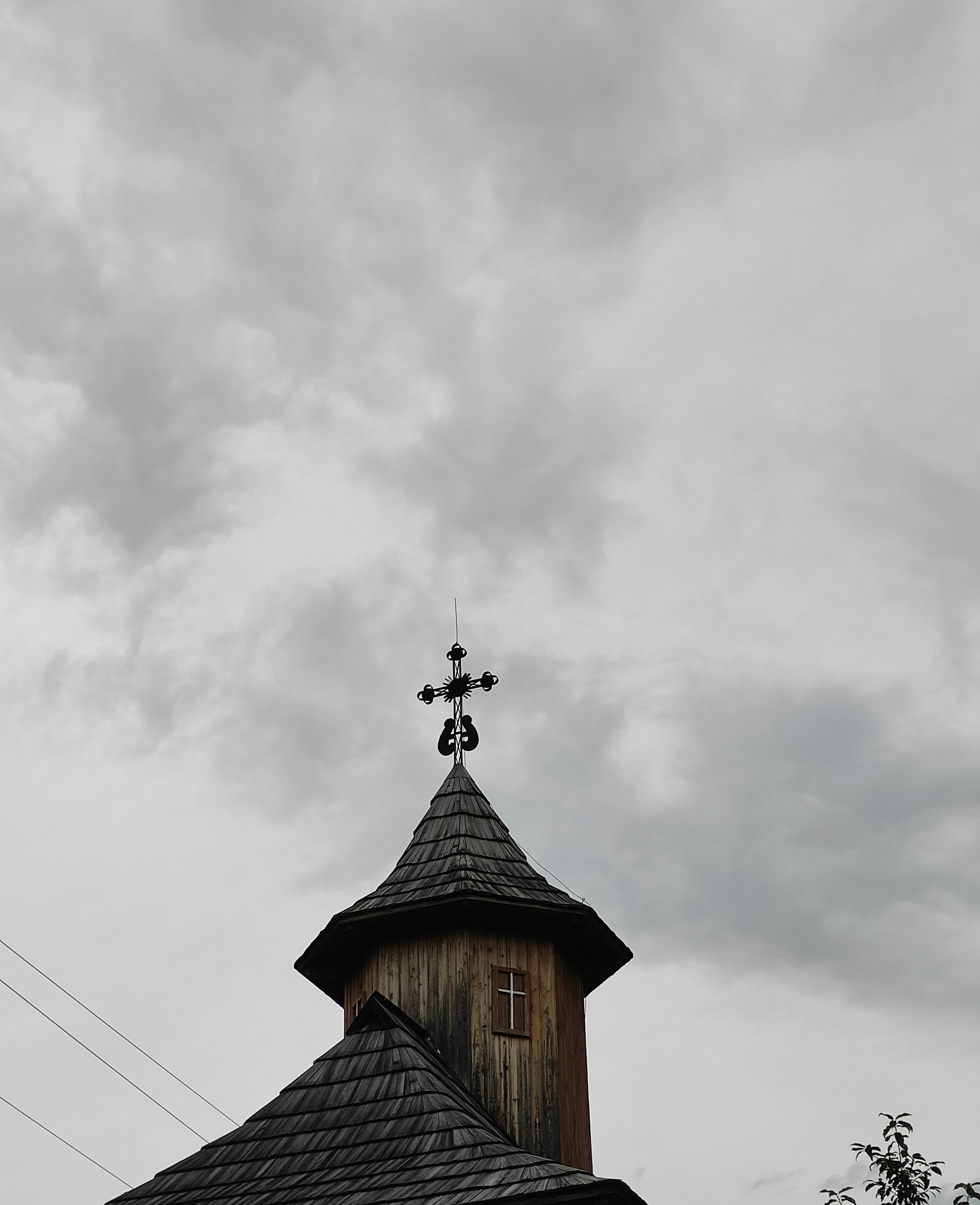Wooden church steeple crowned with a cross, set against a dramatic, cloudy sky. The architecture reflects traditional craftsmanship.