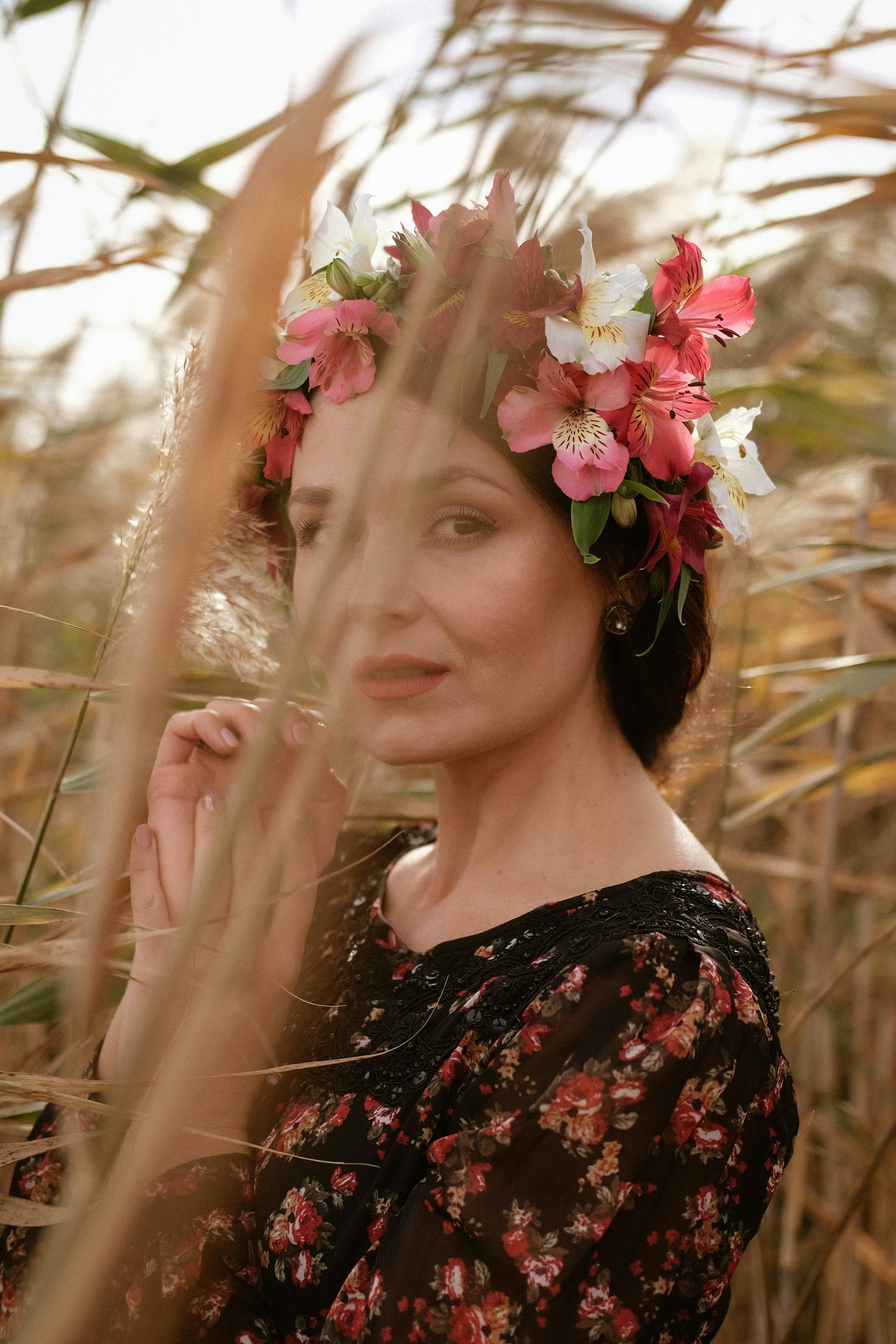 Woman in a black and red floral dress with a pink and white flower headdress, surrounded by tall reeds.
