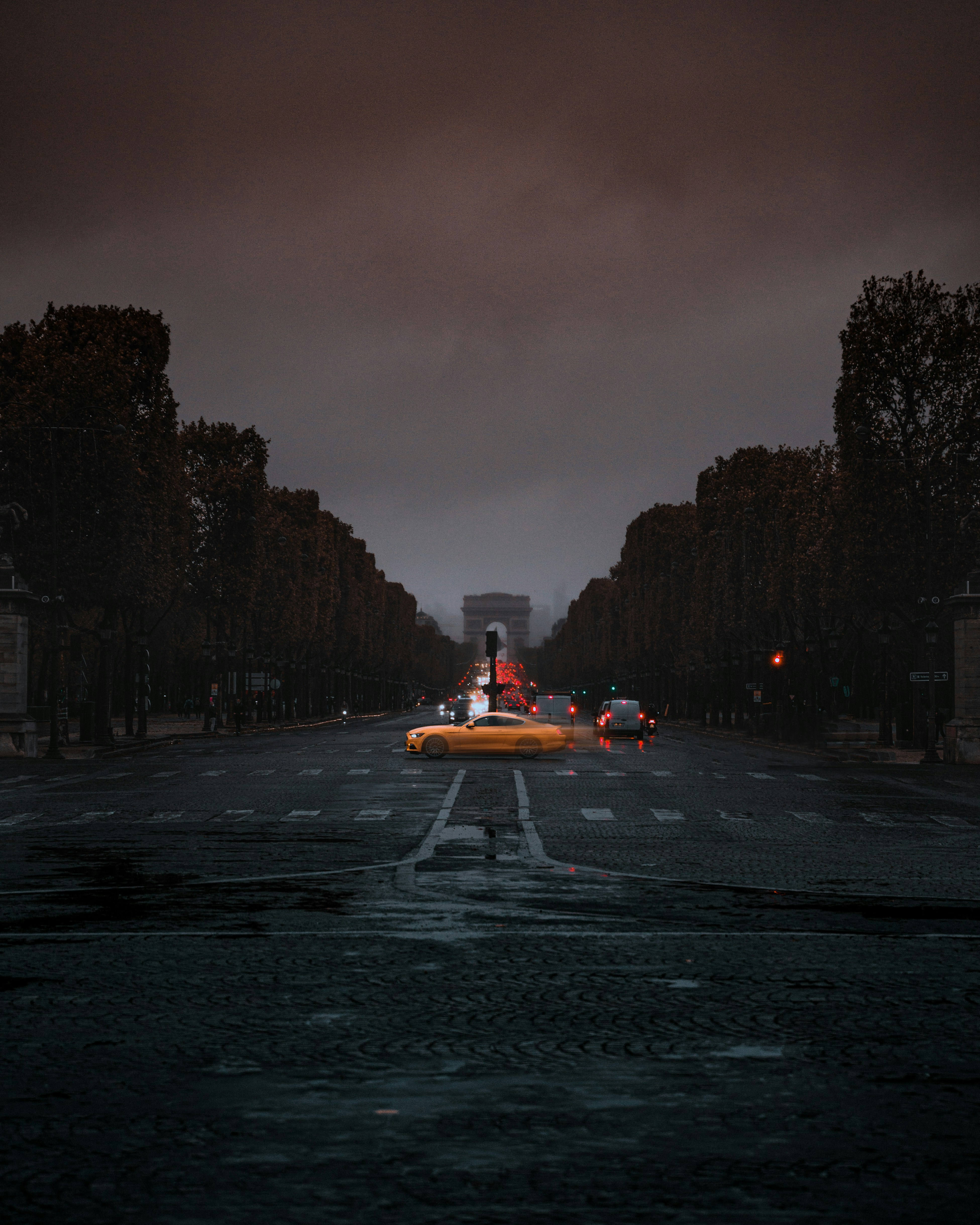A vibrant yellow taxi contrasts against a moody, overcast Parisian street, leading towards the Arc de Triomphe in the distance.