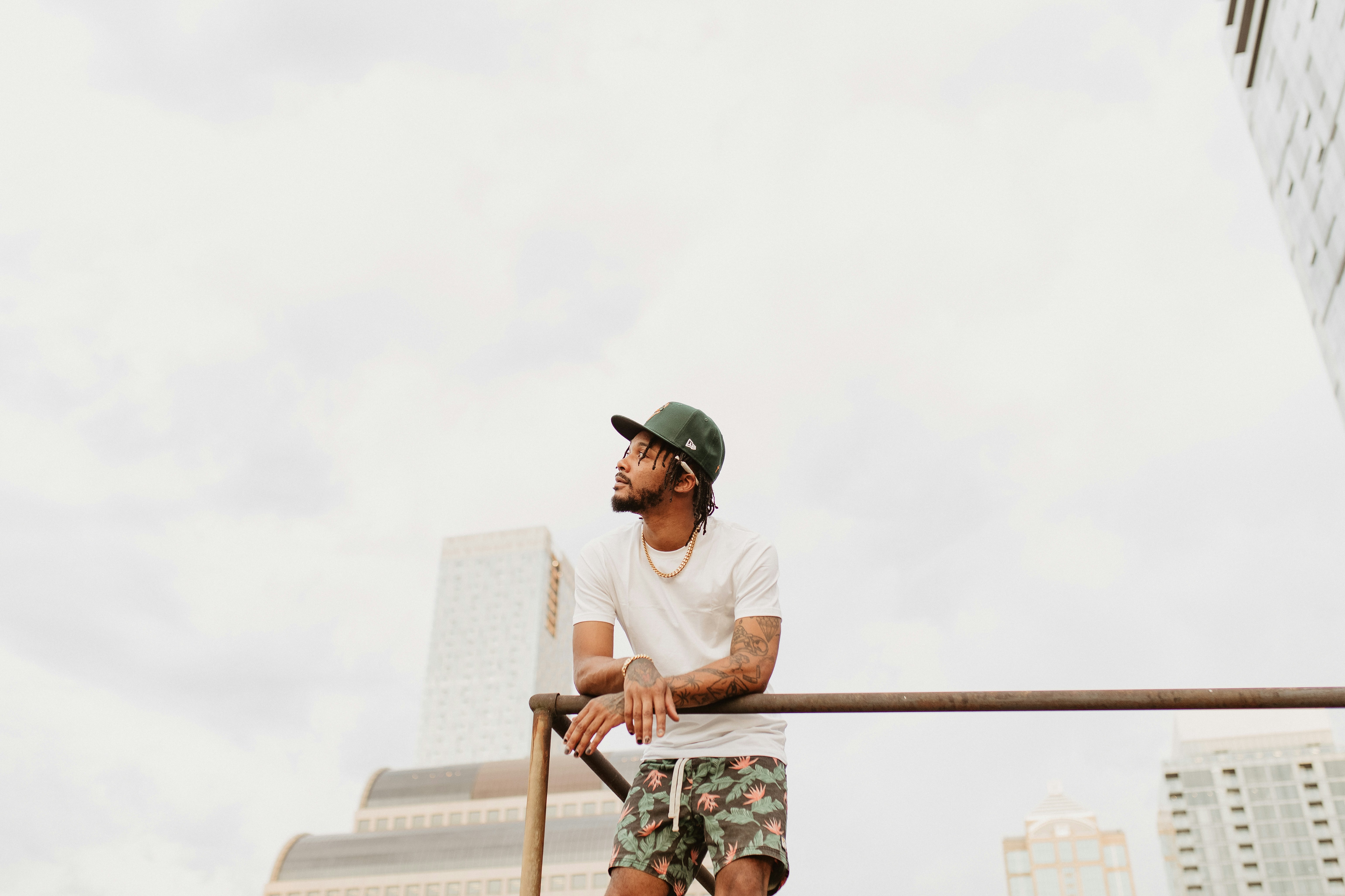 A man leans against a railing, gazing thoughtfully at the skyline, dressed casually in a white t-shirt and camo shorts. The backdrop features towering buildings under a cloudy sky.