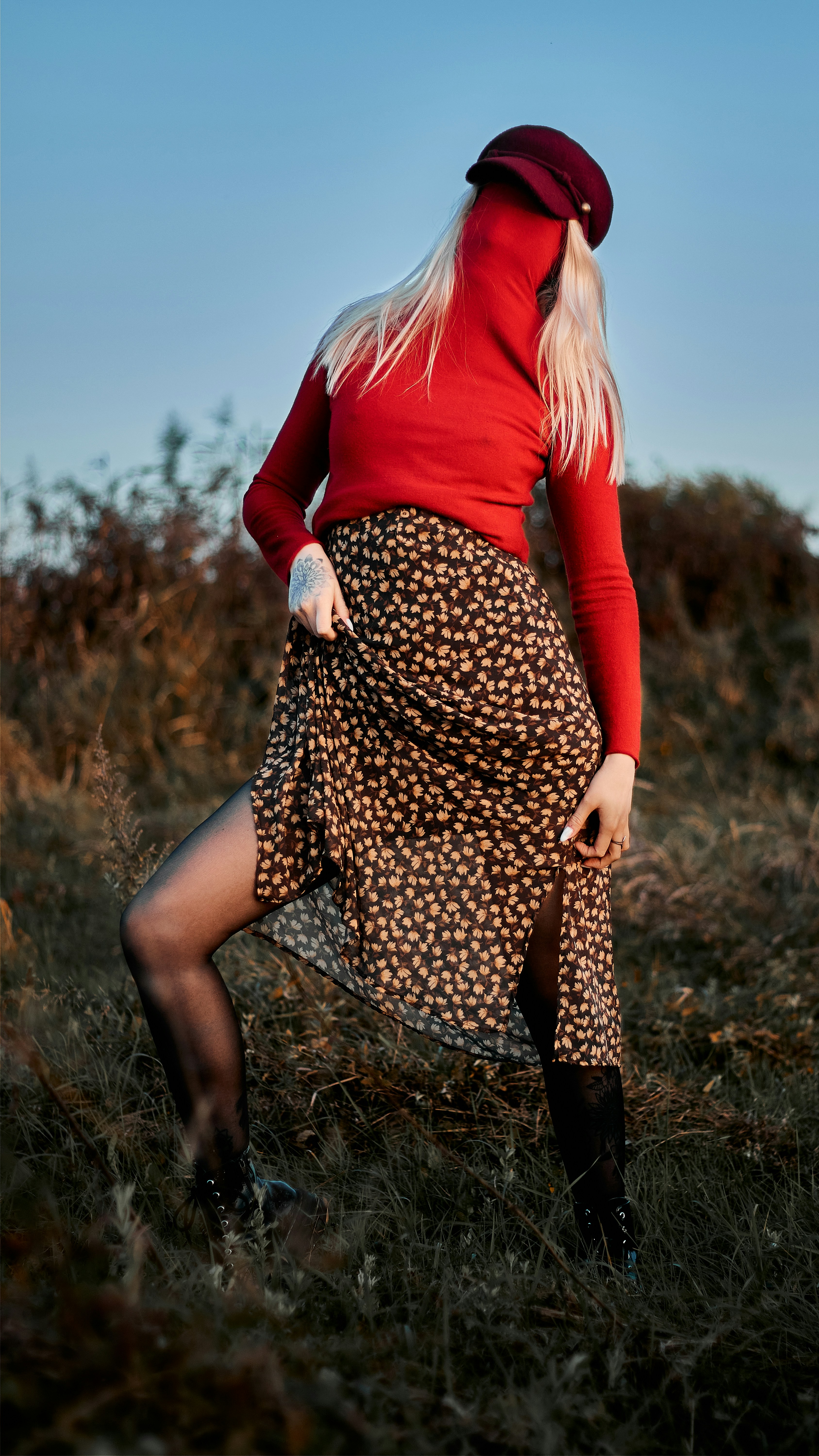 A figure clad in a red top and floral skirt poses playfully in a grassy field, with a unique styling choice that obscures the face. The warm light enhances the scene's artistic flair.