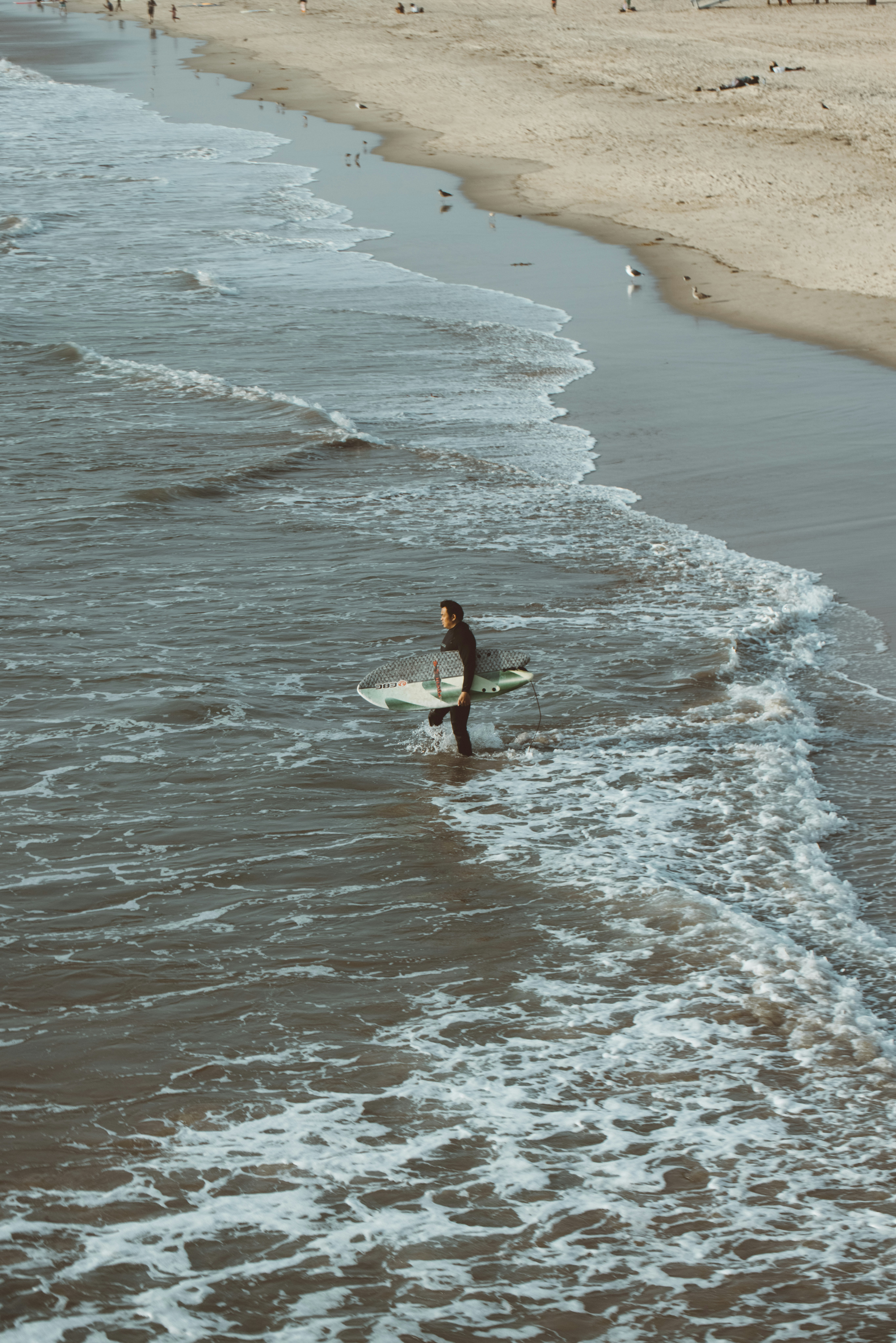 Surfer standing at the water's edge, preparing to ride the waves as the tide rolls in. Soft sunlight reflects off the water's surface.