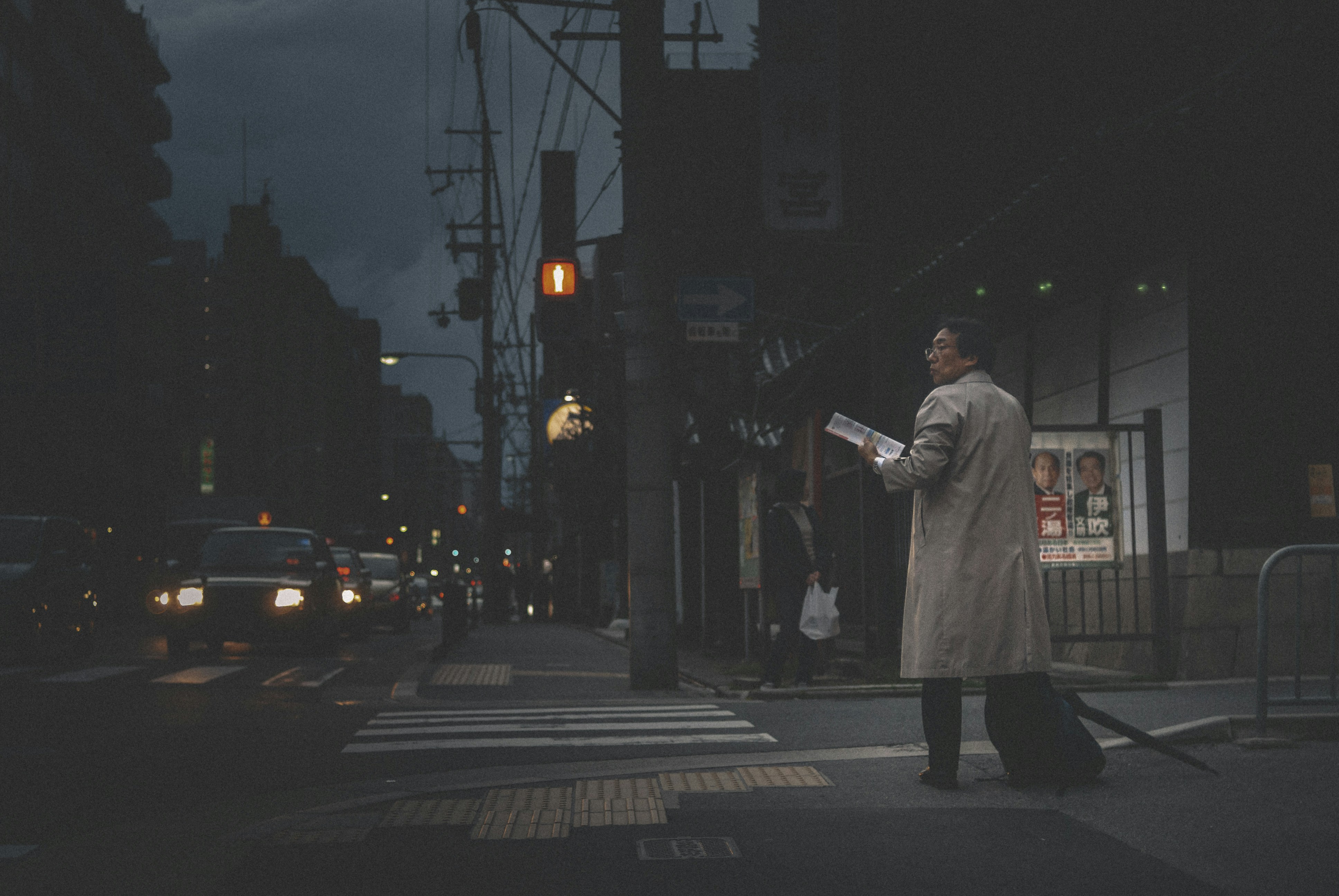 A lone figure in a trench coat stands at a crosswalk, holding a map while illuminated by streetlights in a bustling city environment.