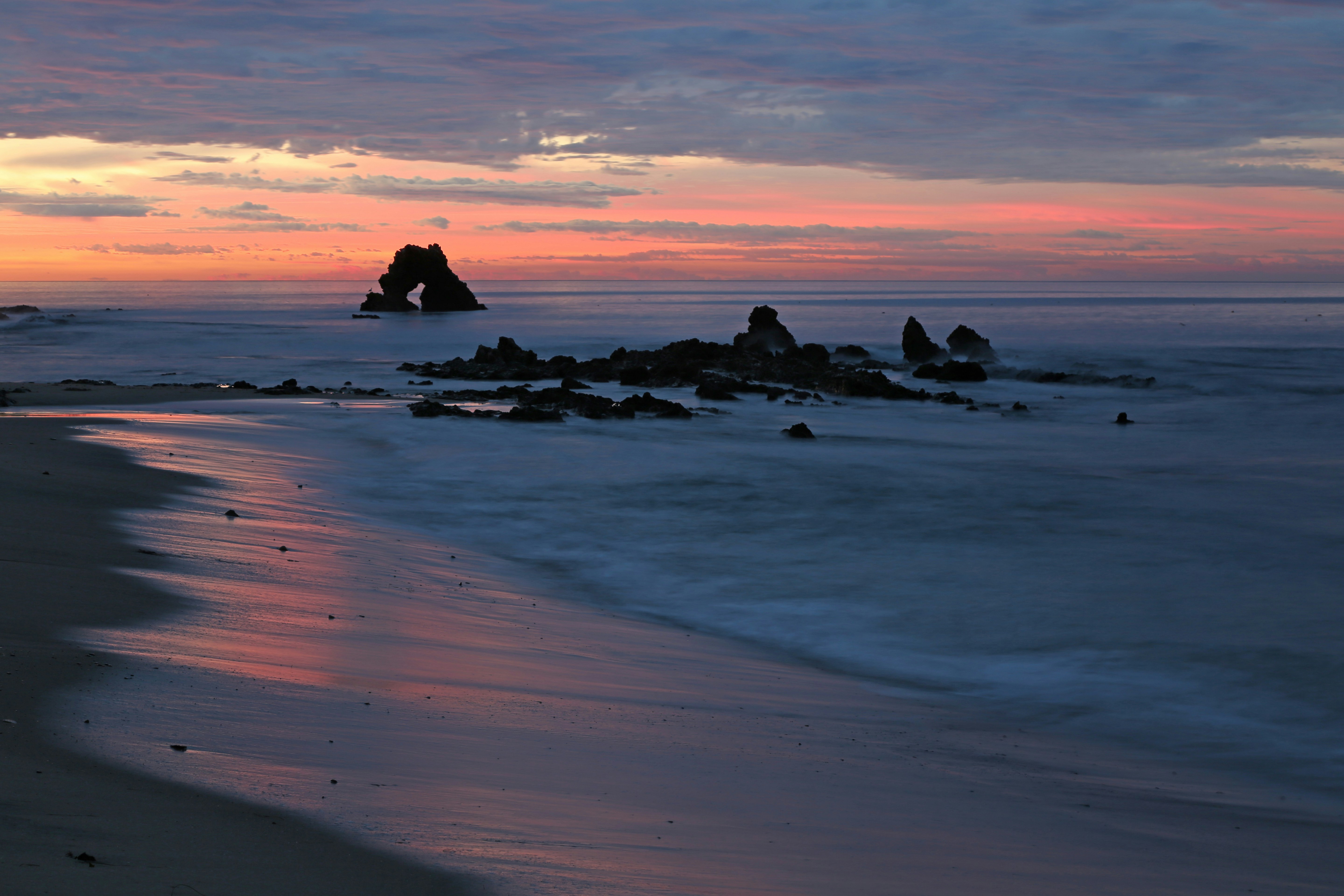 Waves gently lap at the shore under a colorful twilight sky, with rocky formations silhouetted against the horizon.
