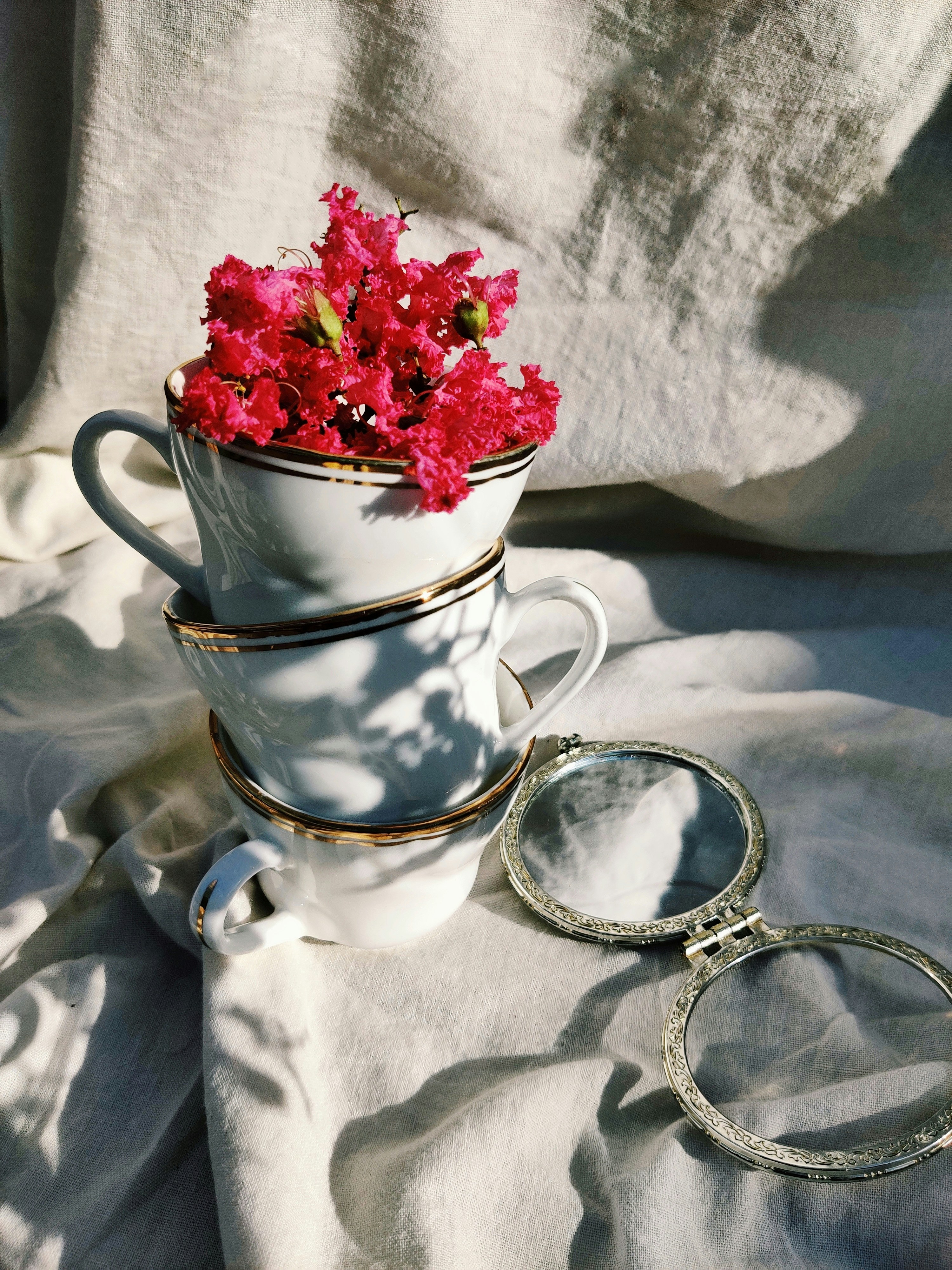 Three stacked white teacups adorned with gold accents, topped with vibrant pink flowers, alongside an open vintage compact mirror on a soft fabric background.