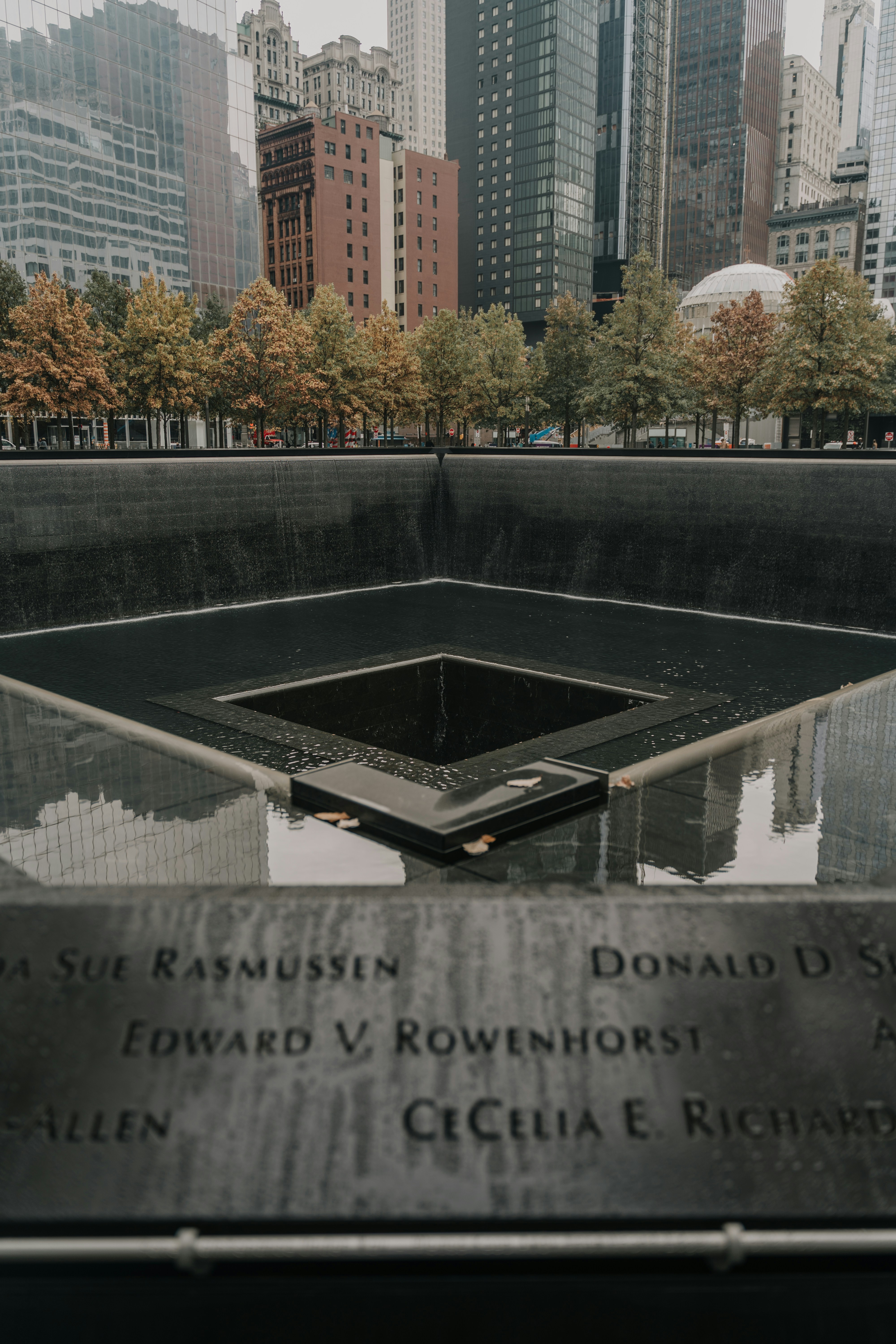 The 9/11 Memorial features a solemn water feature with names inscribed on a dark stone edge, surrounded by trees and skyscrapers. The reflective water symbolizes loss and remembrance.