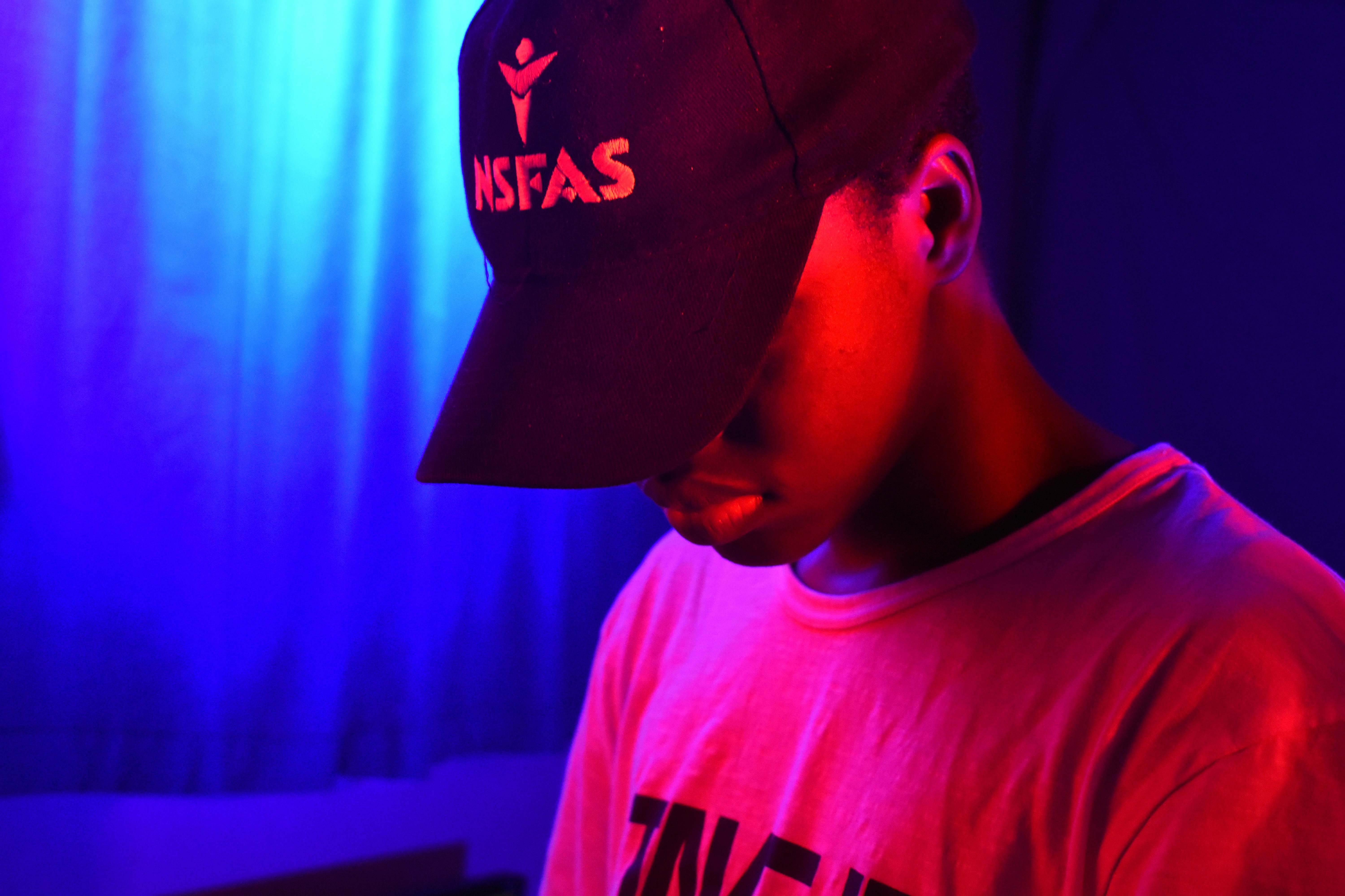 A young individual wearing a cap with the NSFAS logo, bathed in contrasting blue and red lighting, creates a moody atmosphere. The focus is on their introspective expression.