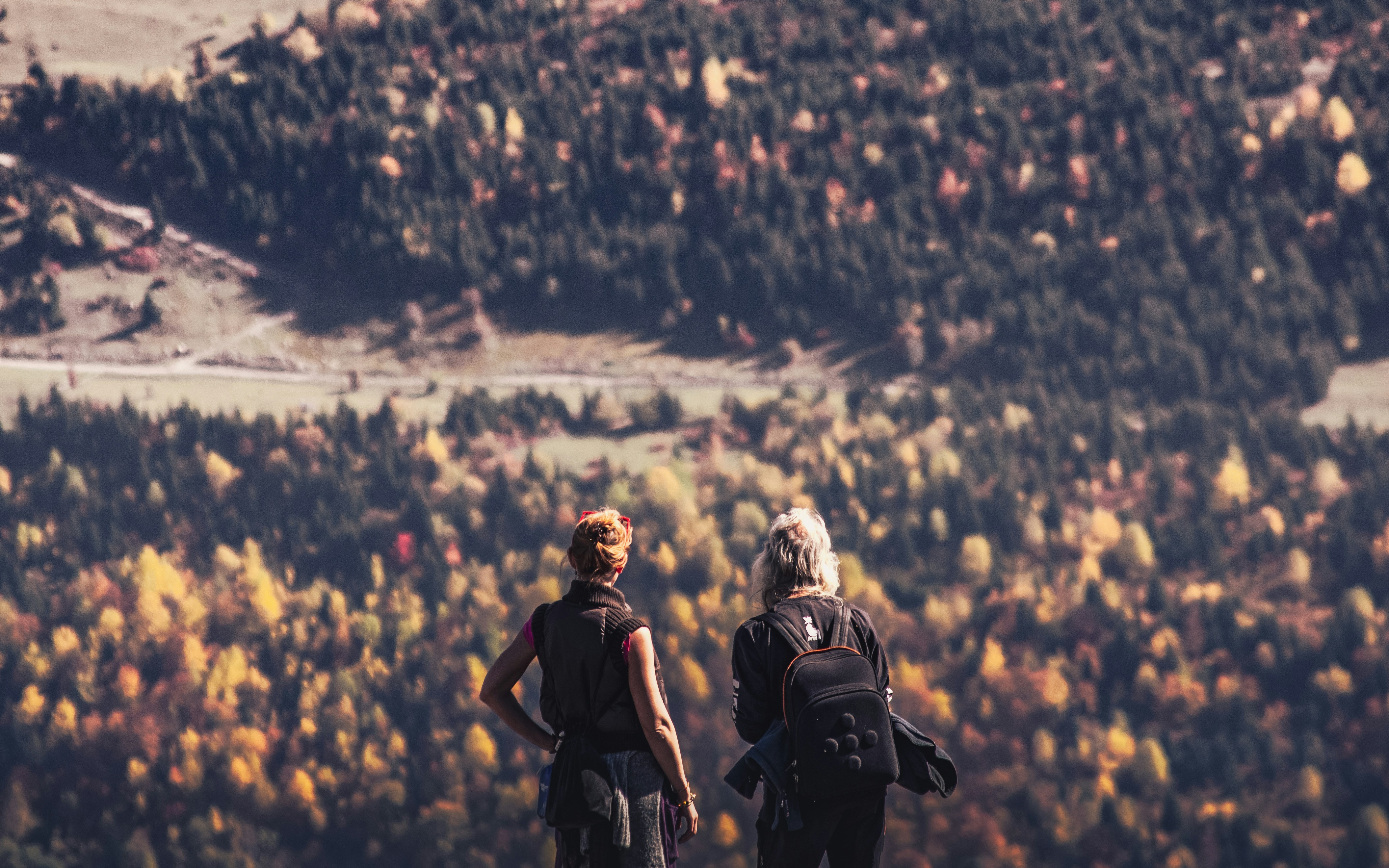 Two hikers stand on a sunlit ridge, overlooking a valley of autumn foliage. The scene captures a quiet moment of exploration framed by a color-saturated landscape.