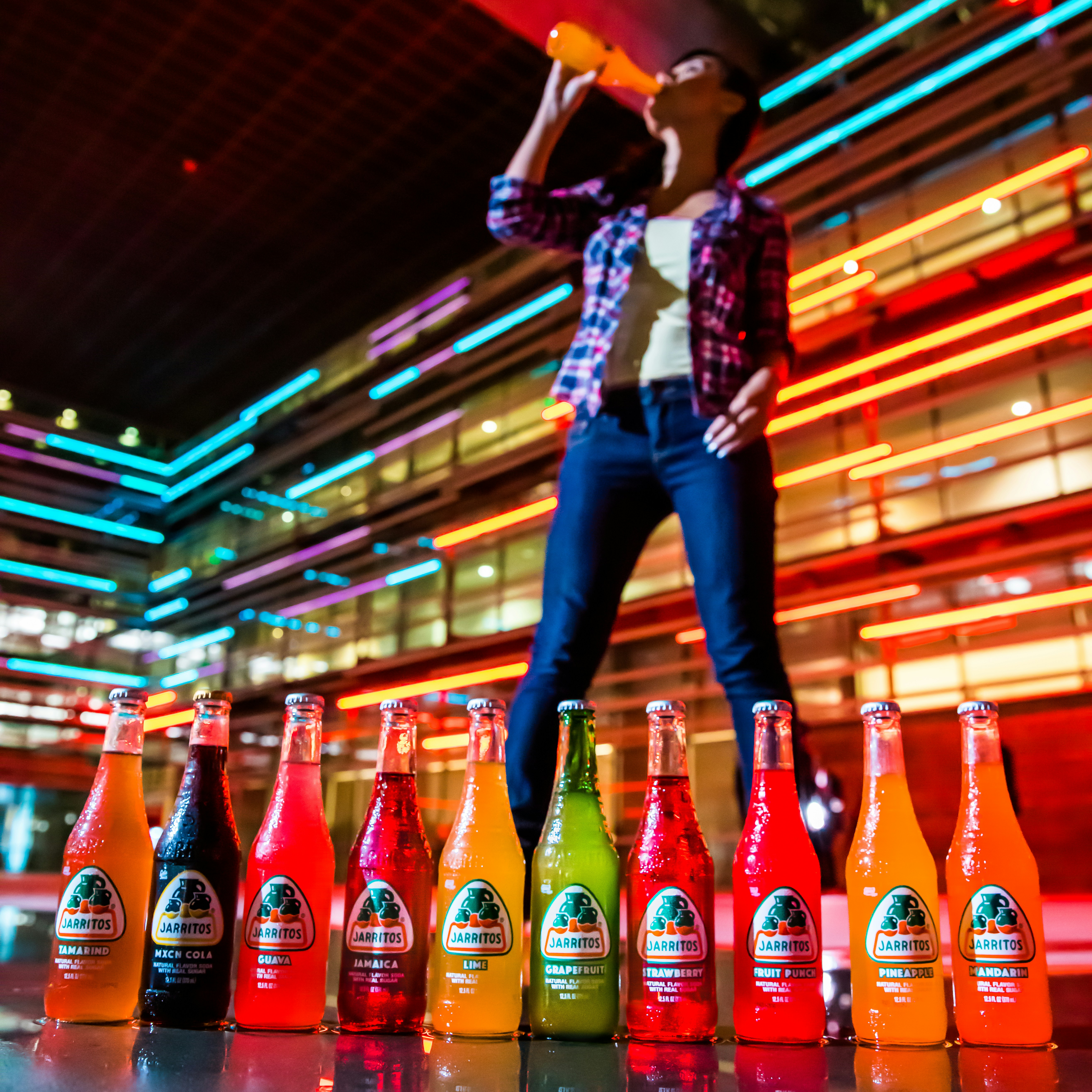 A person enjoying a drink stands amidst a colorful display of Jarritos sodas, illuminated by vibrant neon lights in an urban setting.