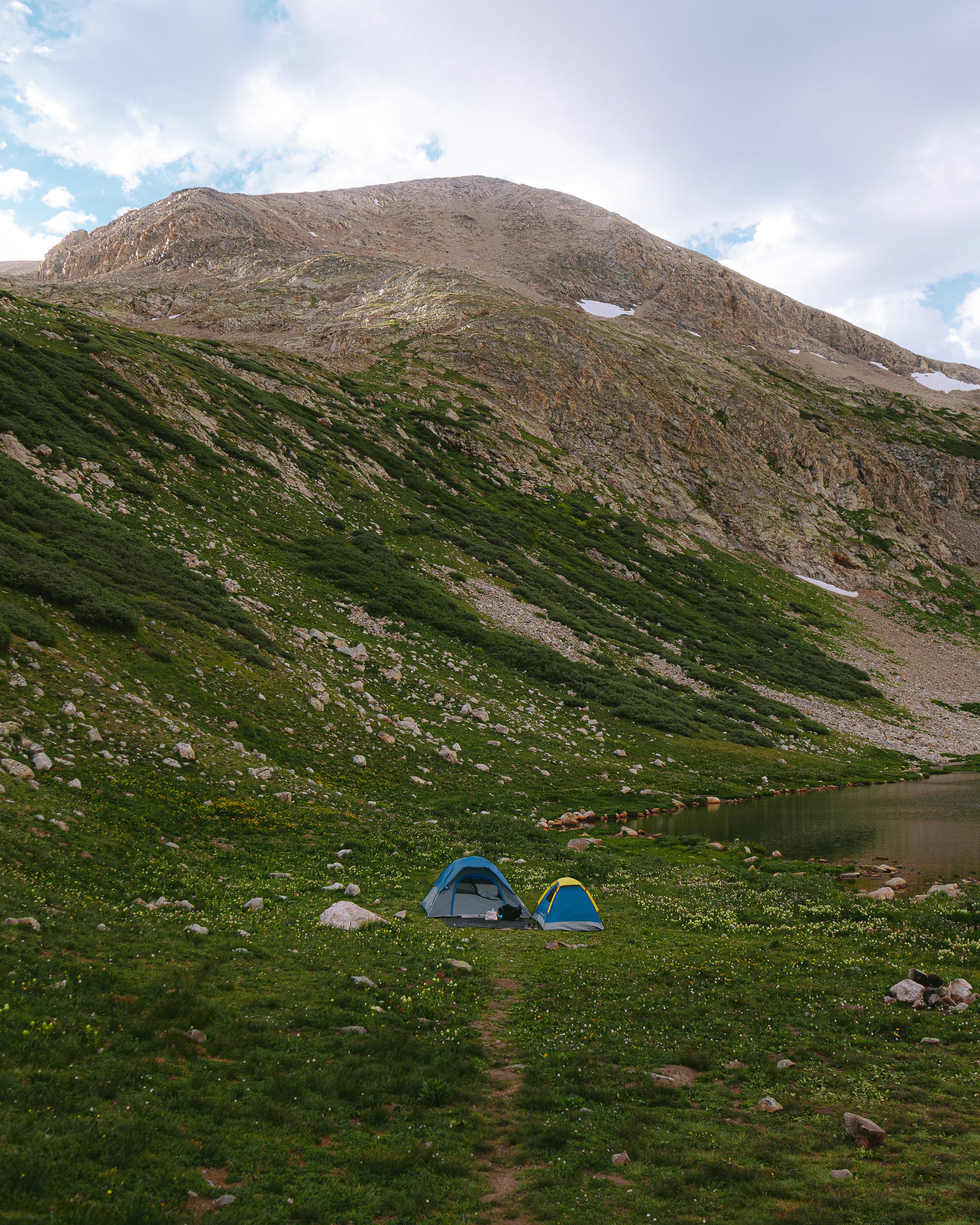 Two colorful tents set against a lush green meadow and a backdrop of towering mountains, reflecting in a tranquil alpine lake.