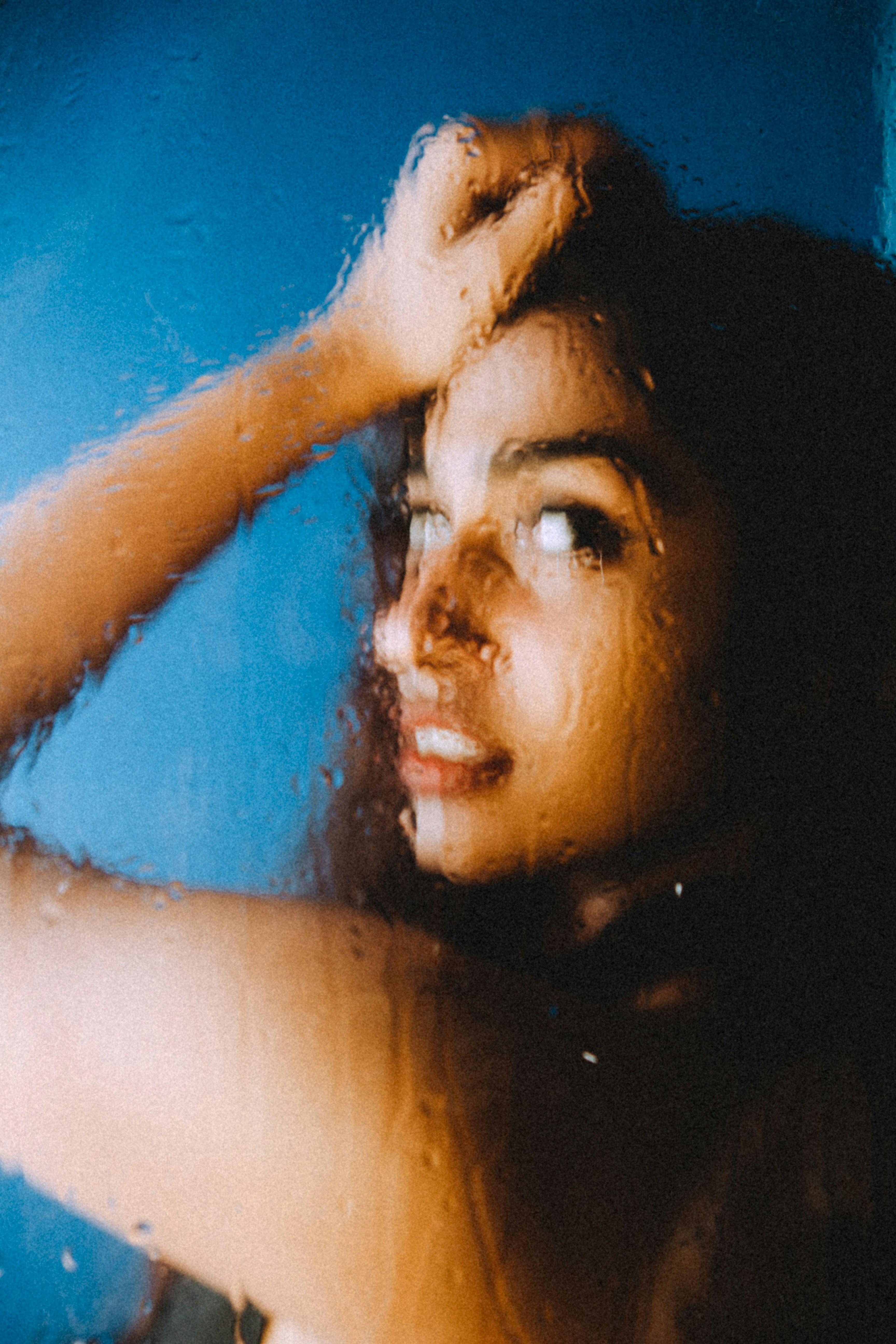 A woman gazes over her shoulder, her features obscured by droplets on a glass surface, creating an ethereal atmosphere. The backdrop is a vibrant blue, enhancing the mood.