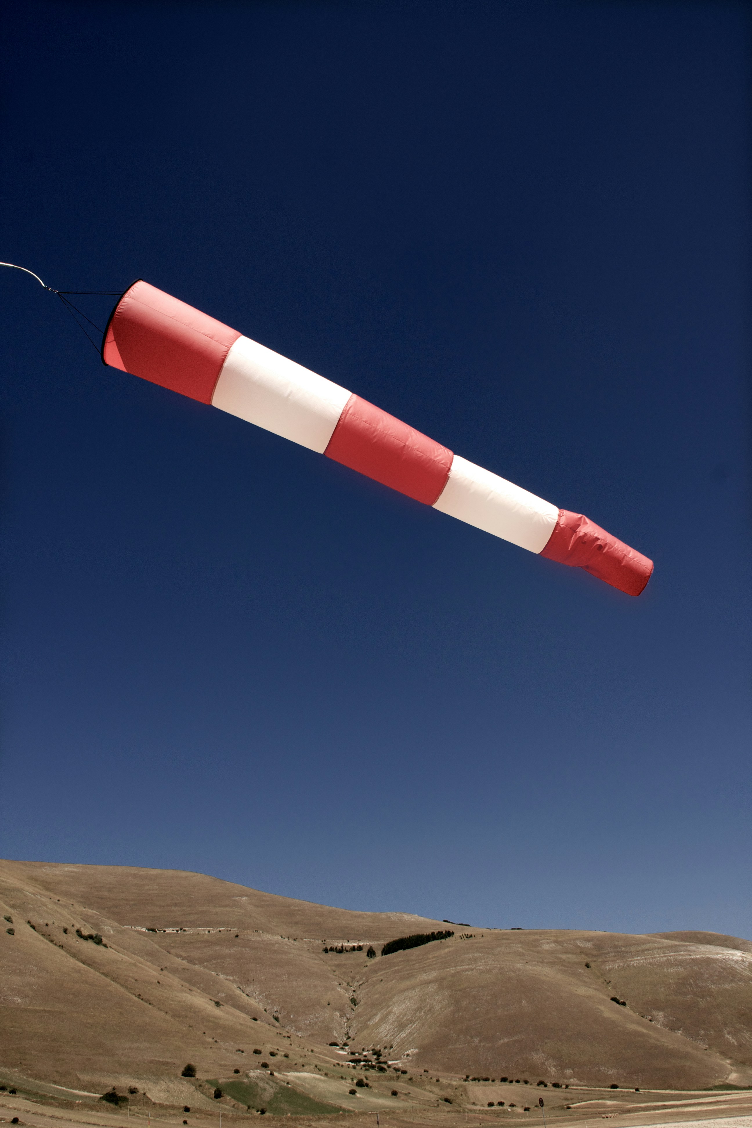 A windsock sways gently against a clear blue sky, indicating wind direction above rolling hills. The vibrant red and white stripes contrast beautifully with the serene landscape.