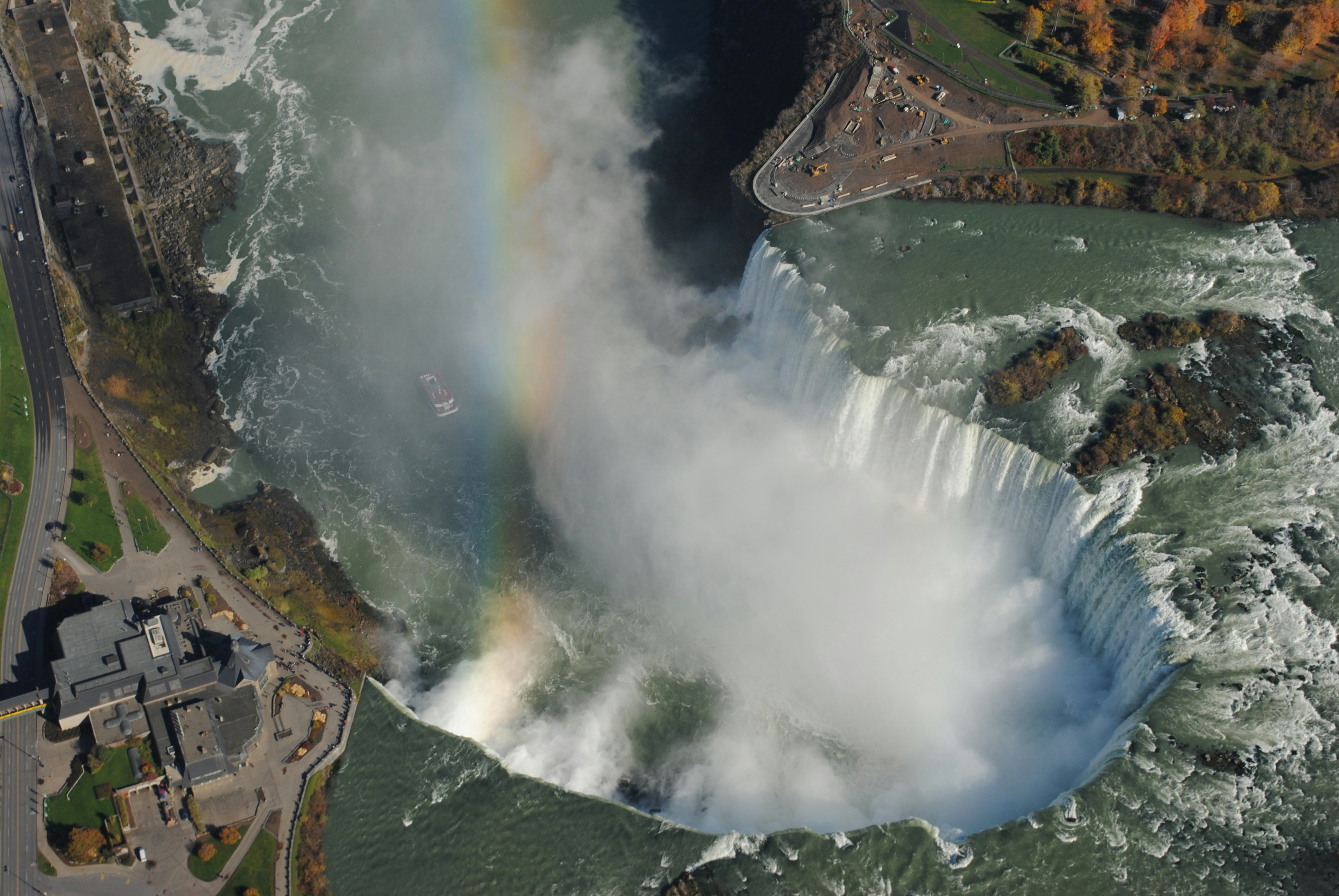 Aerial view of a waterfall with mist creating a vivid rainbow and surrounding autumn foliage.
