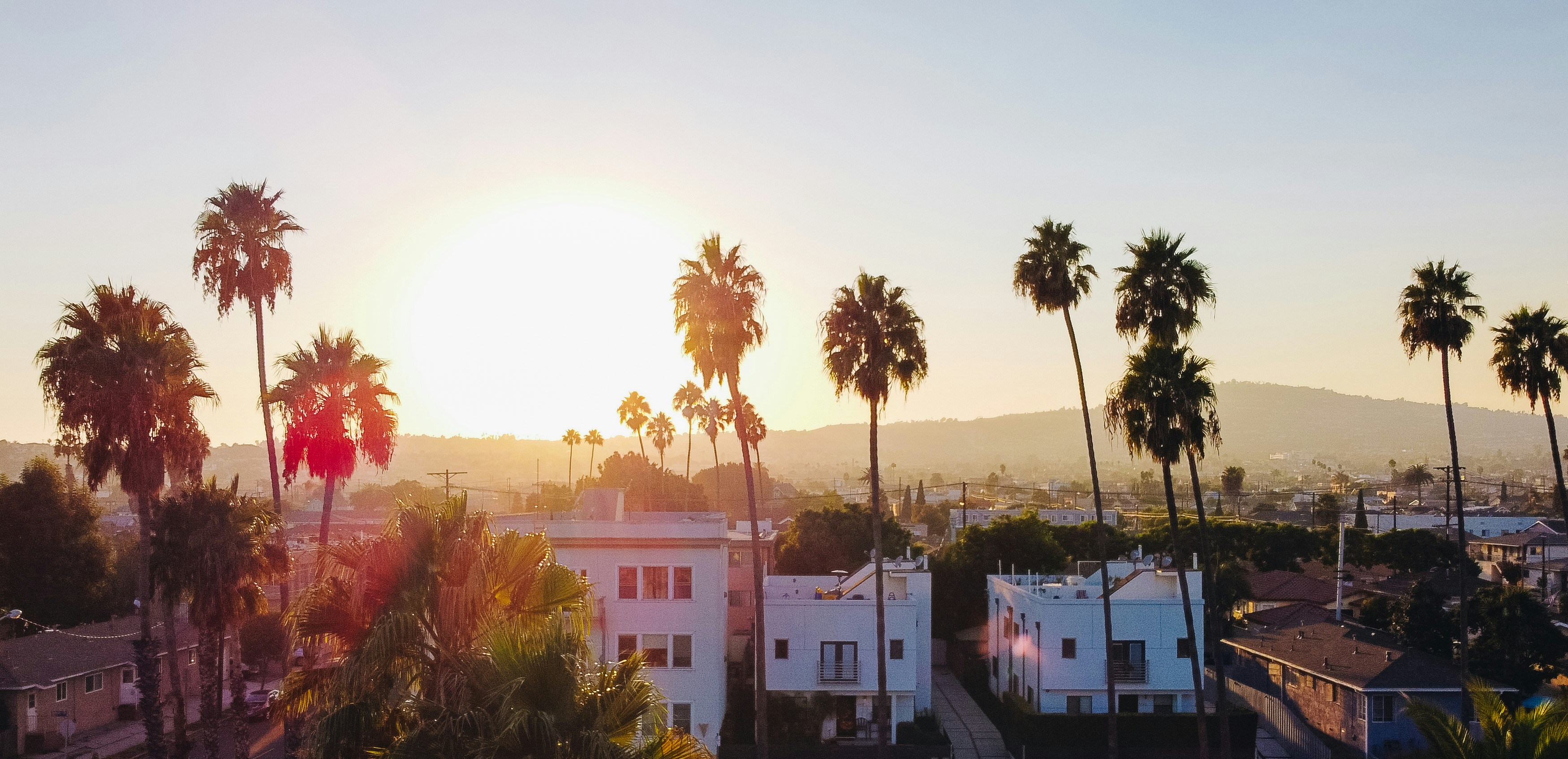 Sunset casting warm hues over palm trees and urban buildings with distant hills.