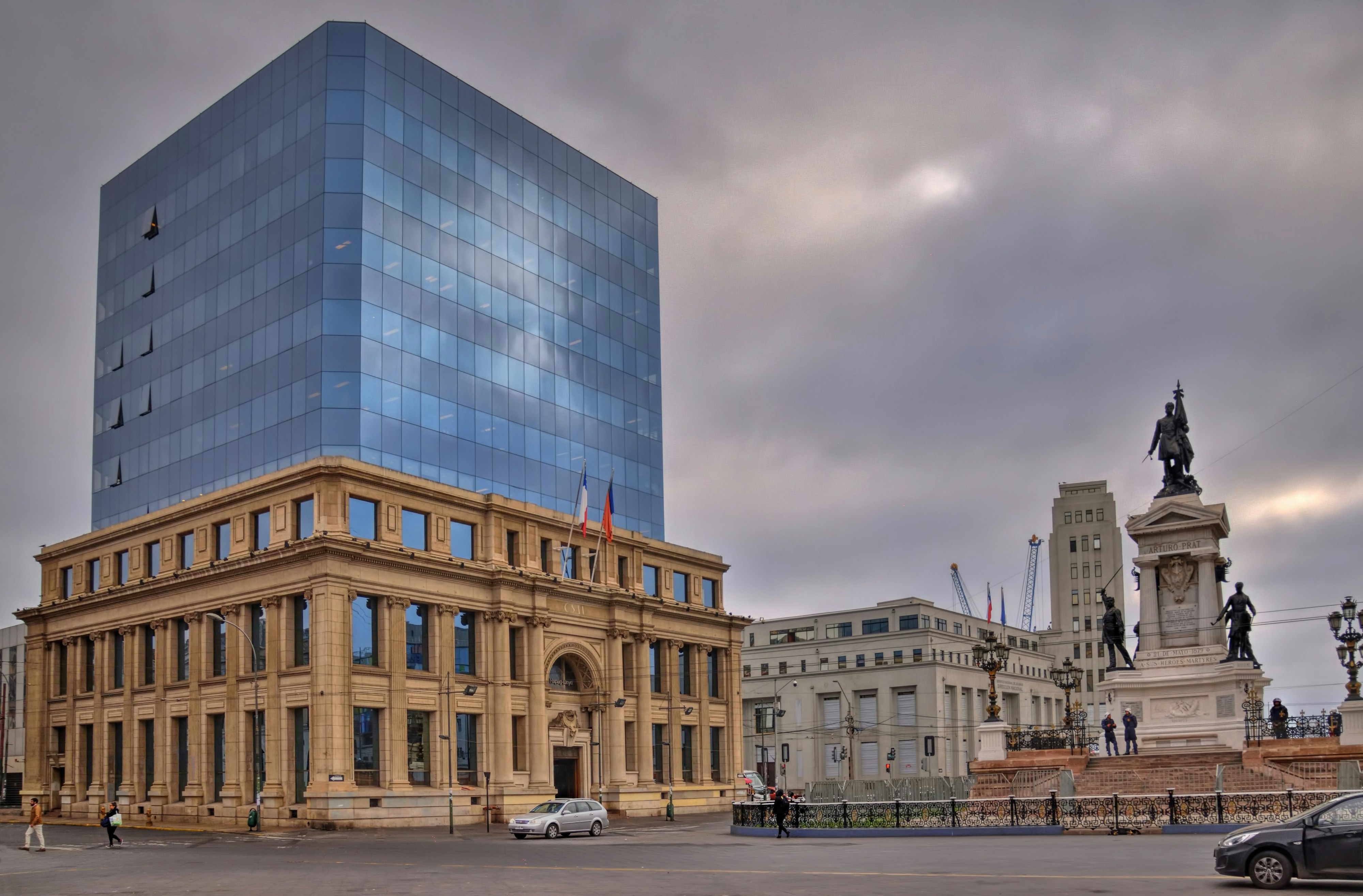 Glass-fronted building juxtaposed with a historic monument under a cloudy sky in Valparaiso, Chile.