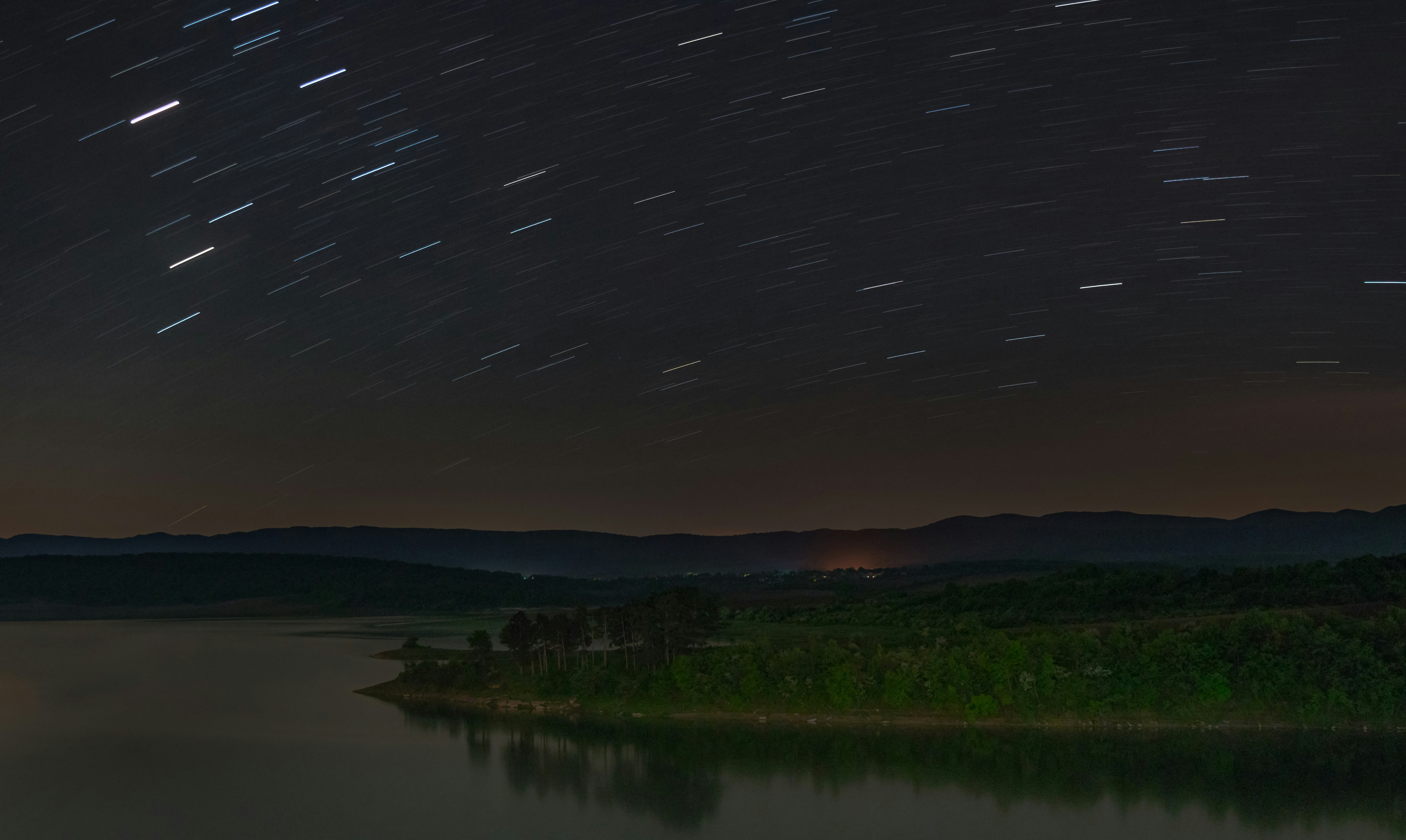 Star trails streak across the night sky above a serene lake and lush greenery.
