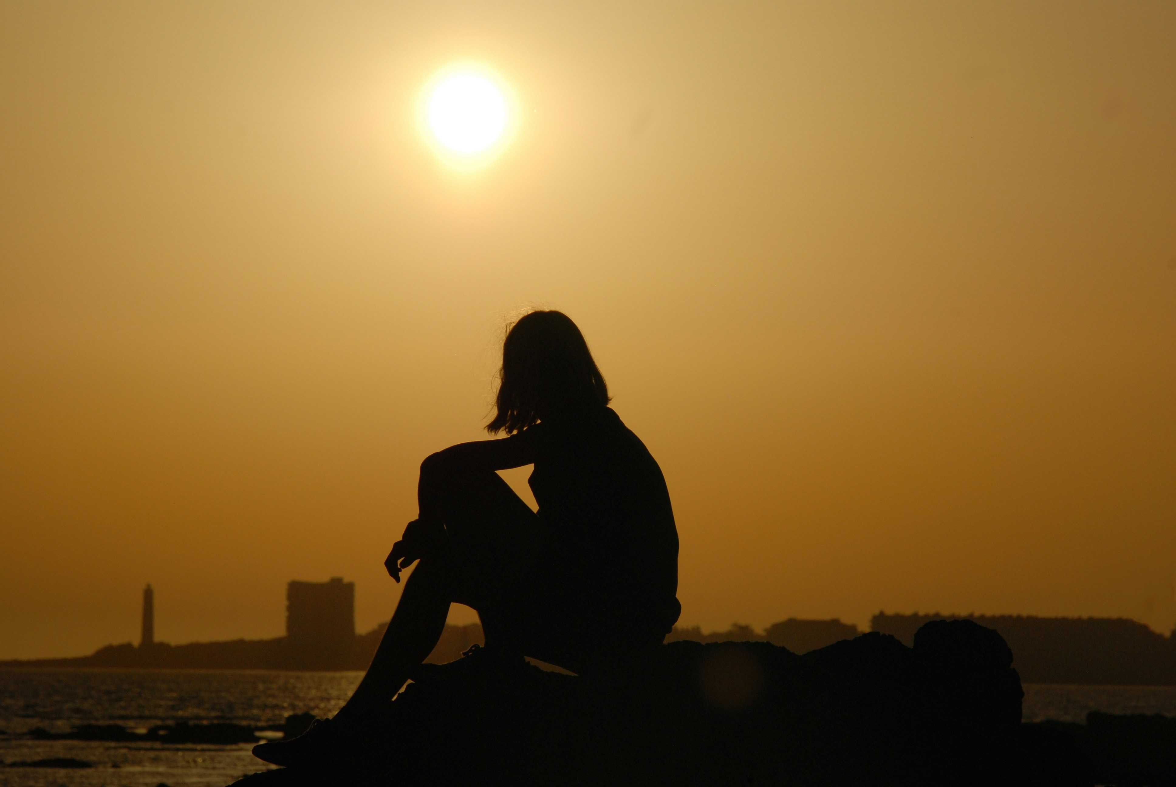Silhouette of a person seated on a rock against a golden sunset, with a distant lighthouse and buildings in the background.
