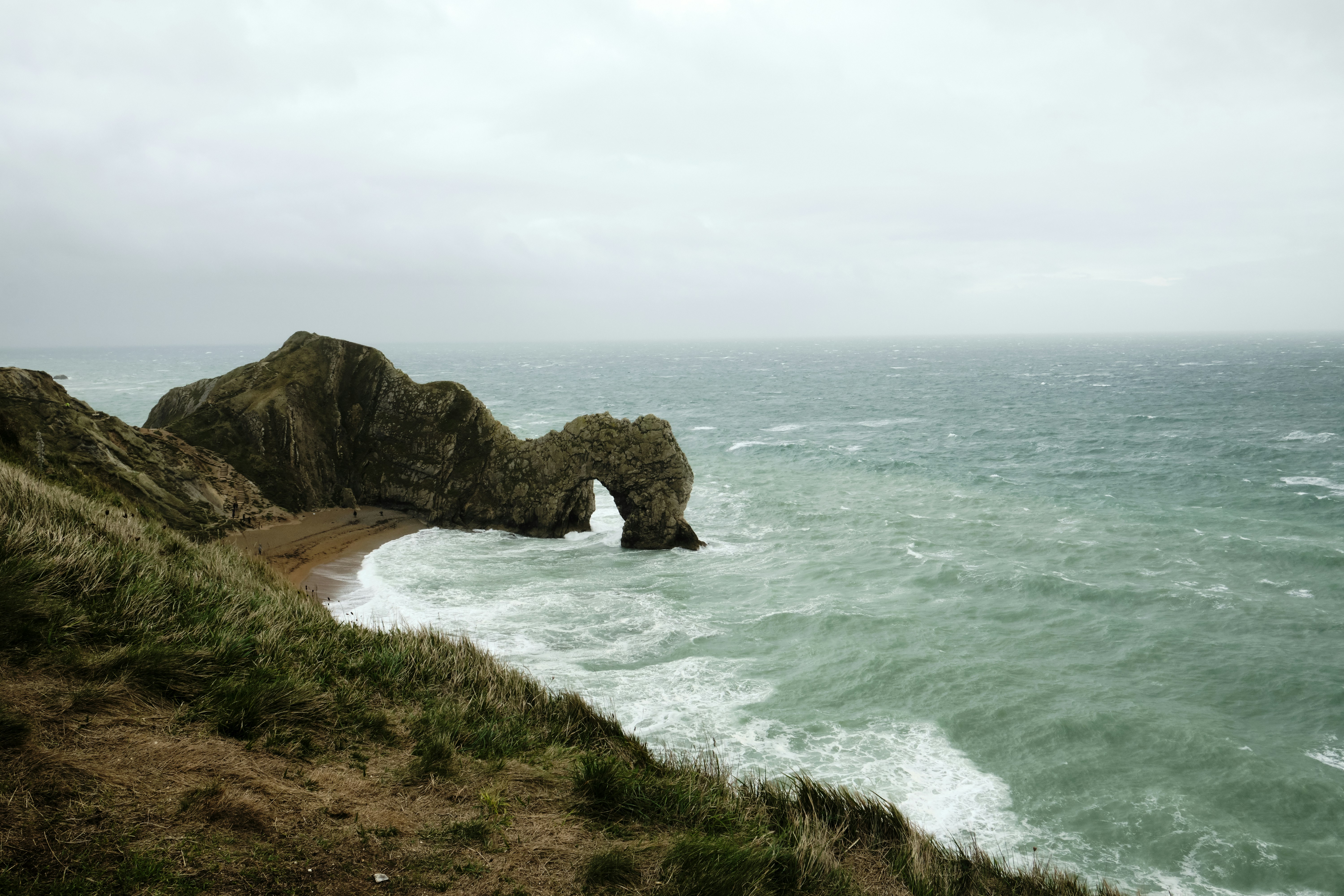 Rocky arch formation rising from the turbulent sea, framed by grassy cliffs under a moody sky. Waves crash against the shore.