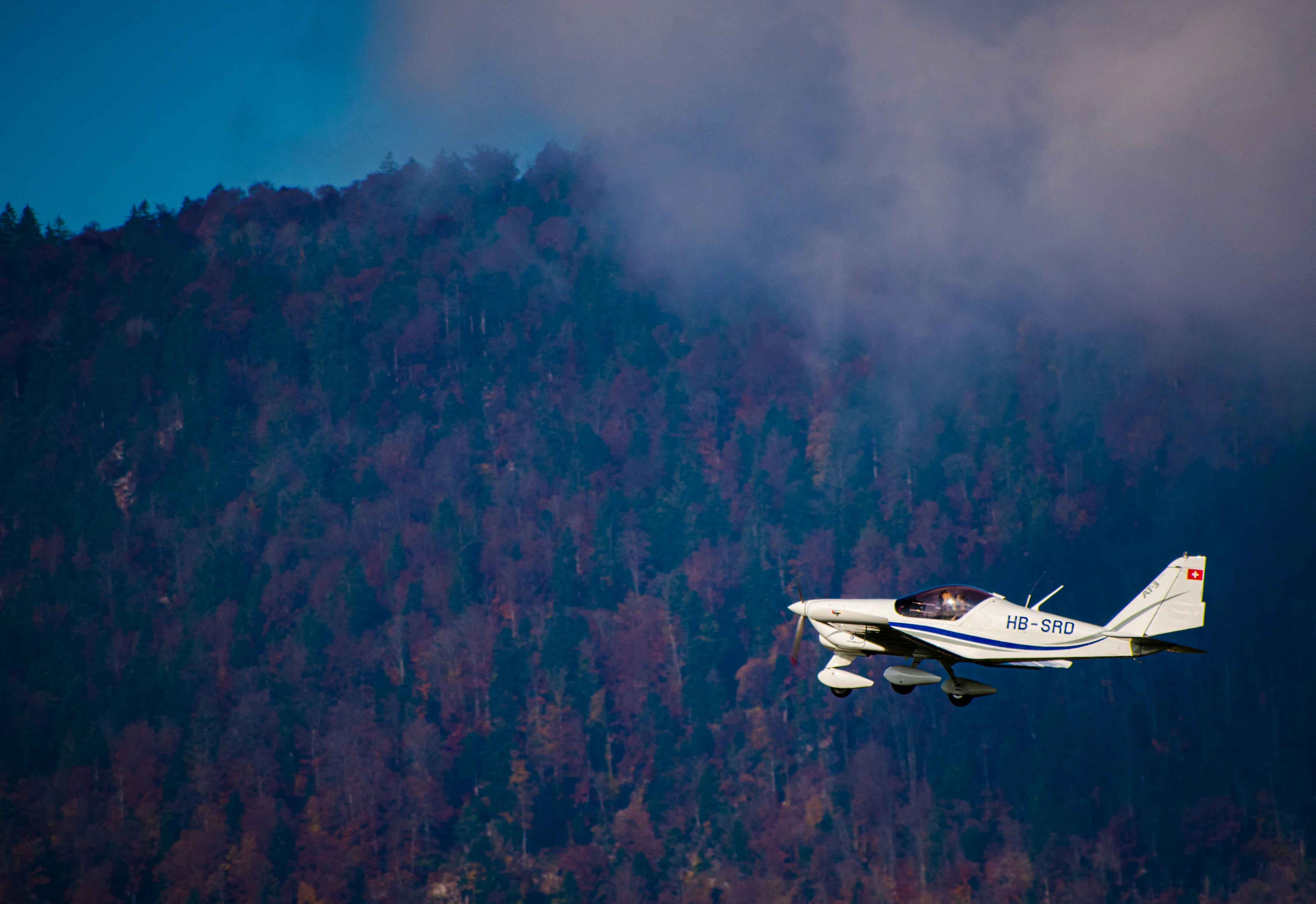 Light aircraft flying against a backdrop of colorful autumn foliage and dramatic clouds.