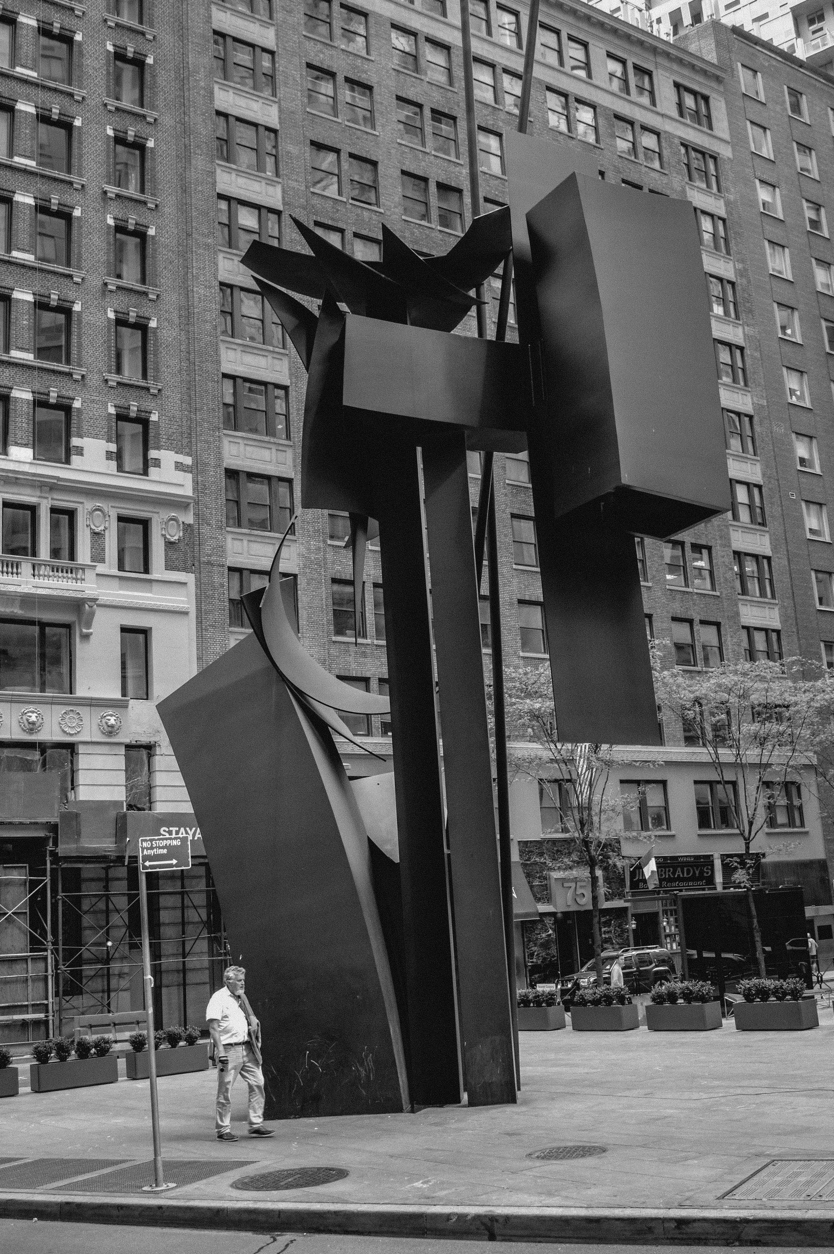 Abstract black sculpture towering over a pedestrian in an urban setting, highlighting the interplay of light and form.