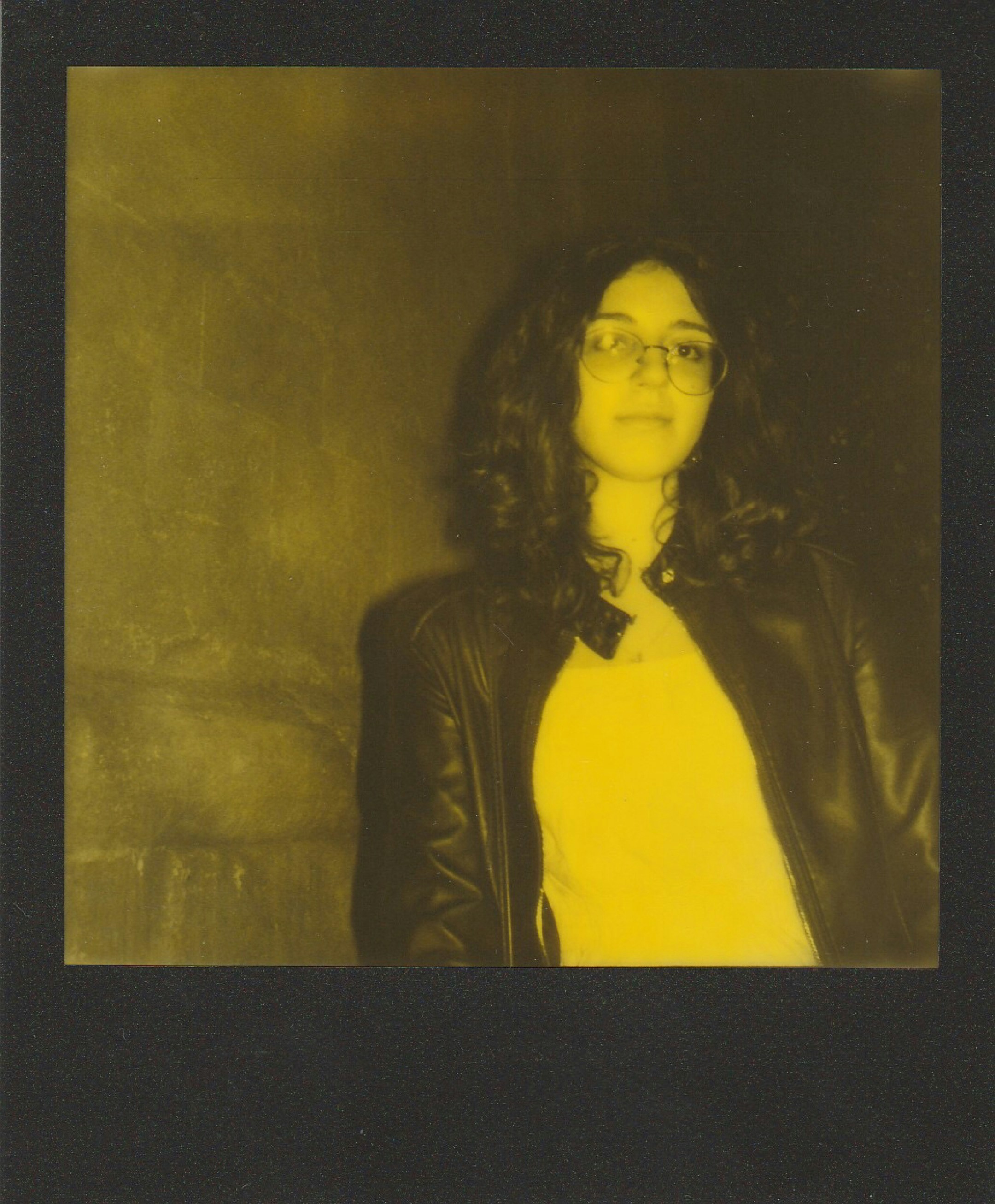 A young woman with curly hair and glasses stands against a textured wall, illuminated in warm yellow tones. The scene captures a moment of introspection.