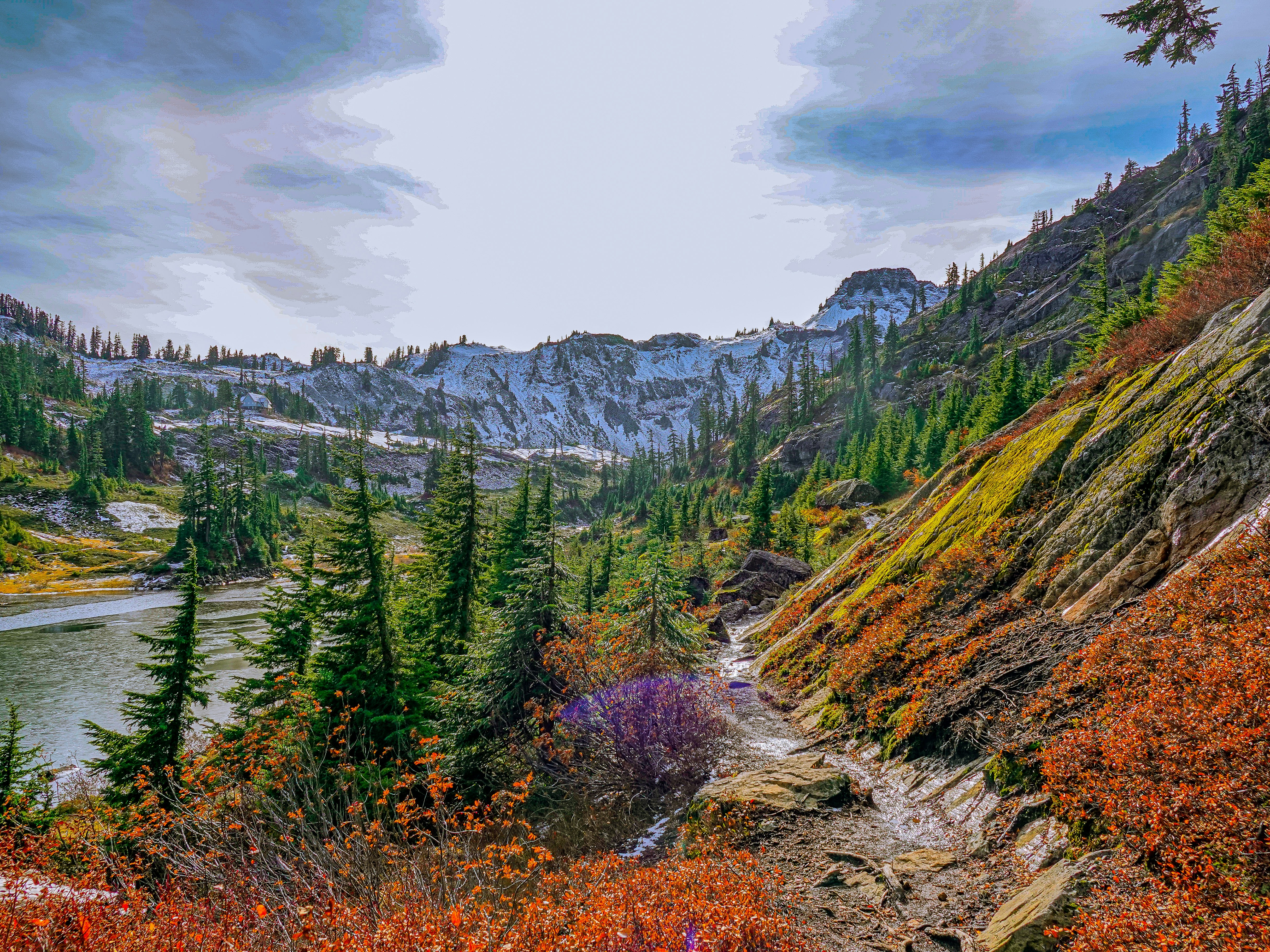Mountain landscape with snow-dusted peaks, evergreen trees, and a winding river under a dynamic sky.