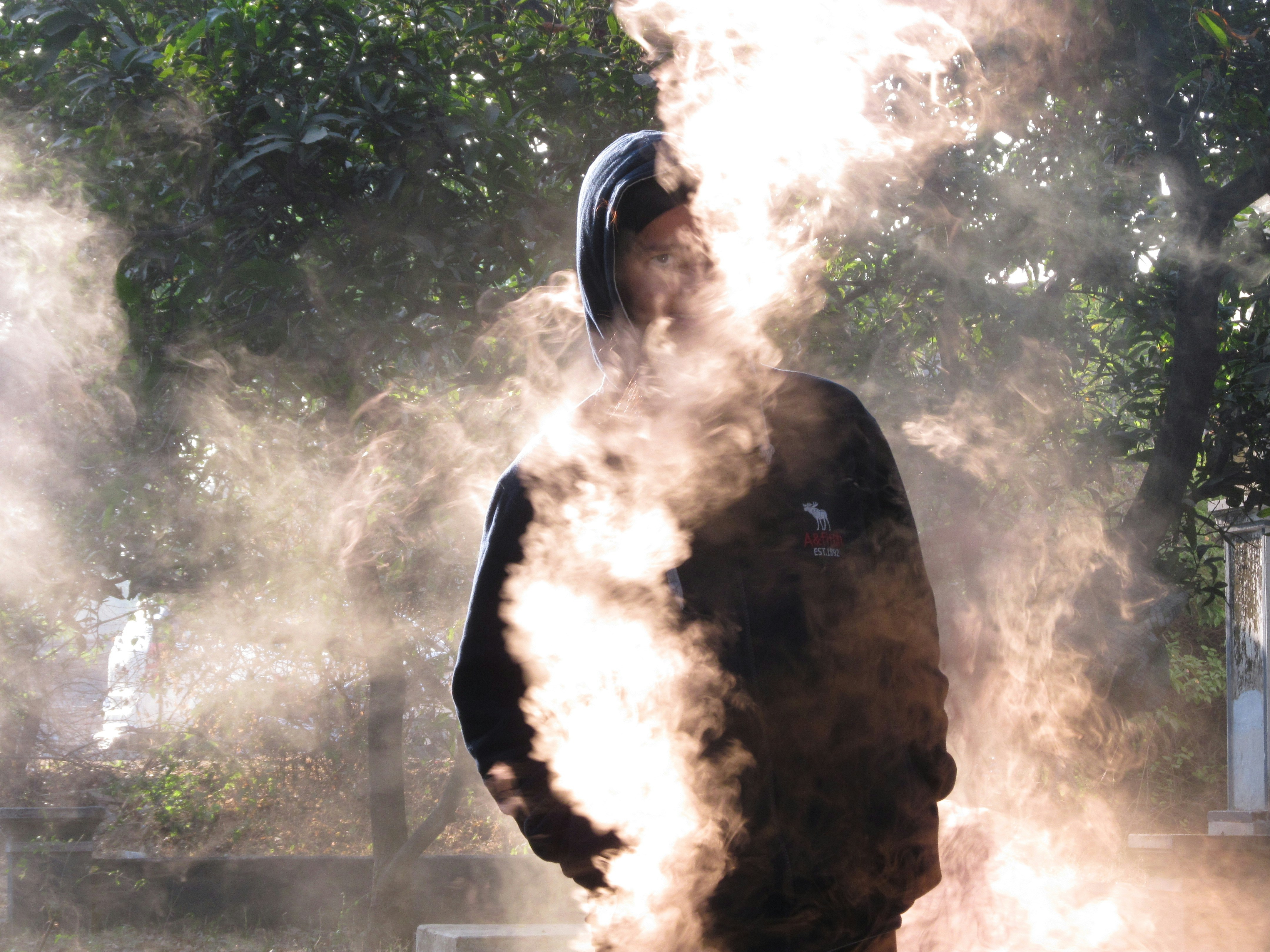Person in a hoodie enveloped in swirling smoke with sunlight filtering through.