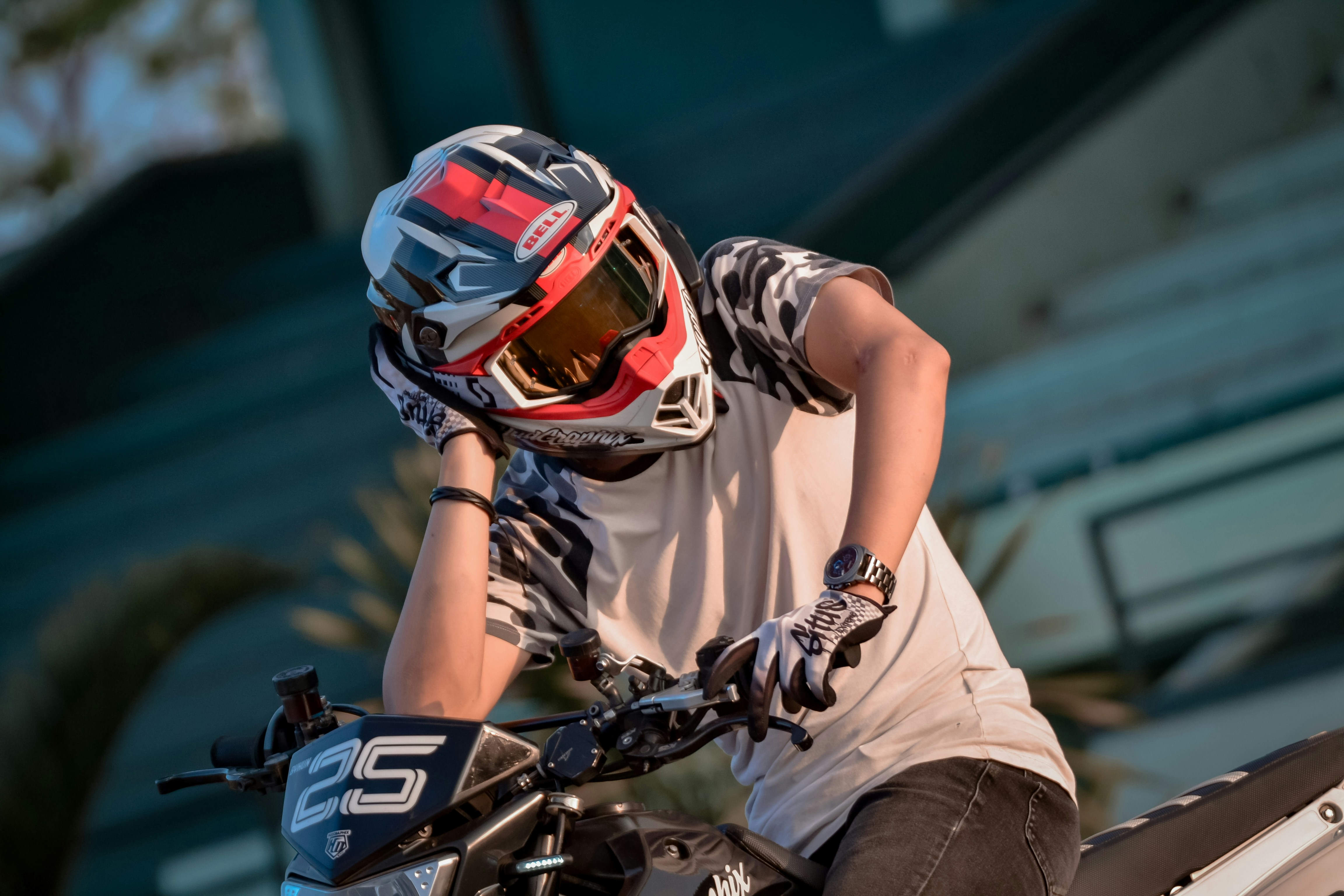 Motocross rider in a helmet poses on a dirt bike with a modern building in the background.