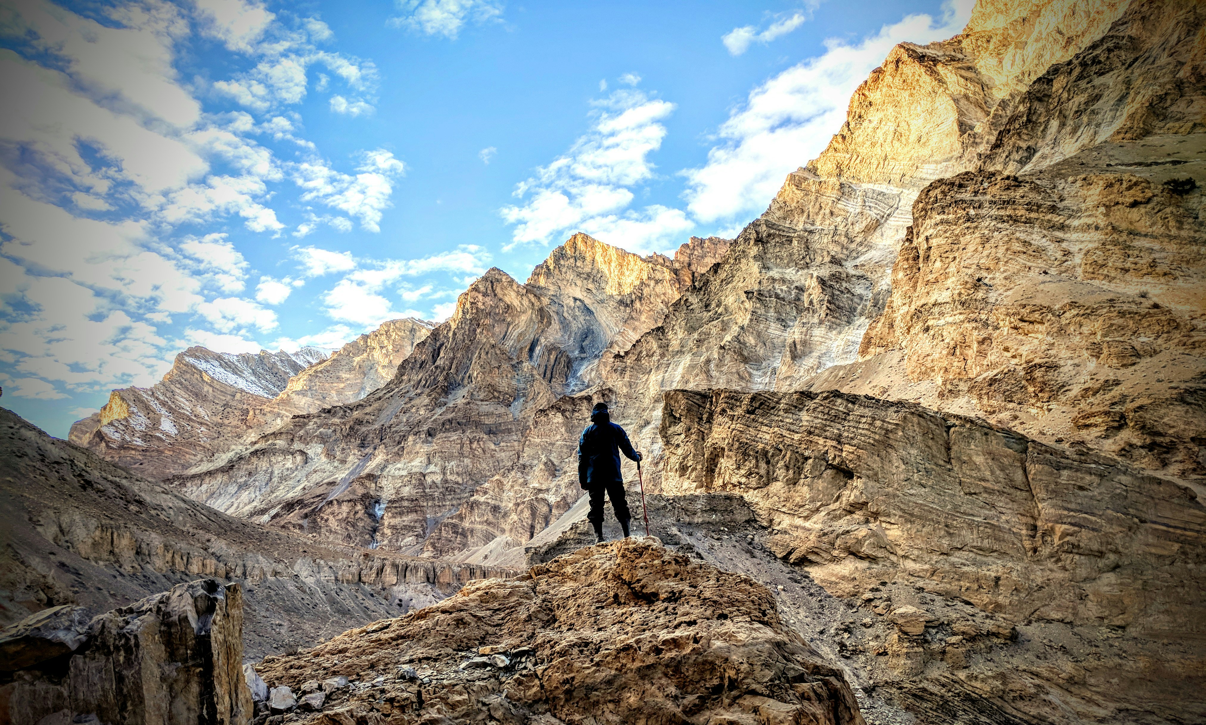 Silhouetted figure stands atop a rocky outcrop, surrounded by towering mountain ranges under a dynamic sky.