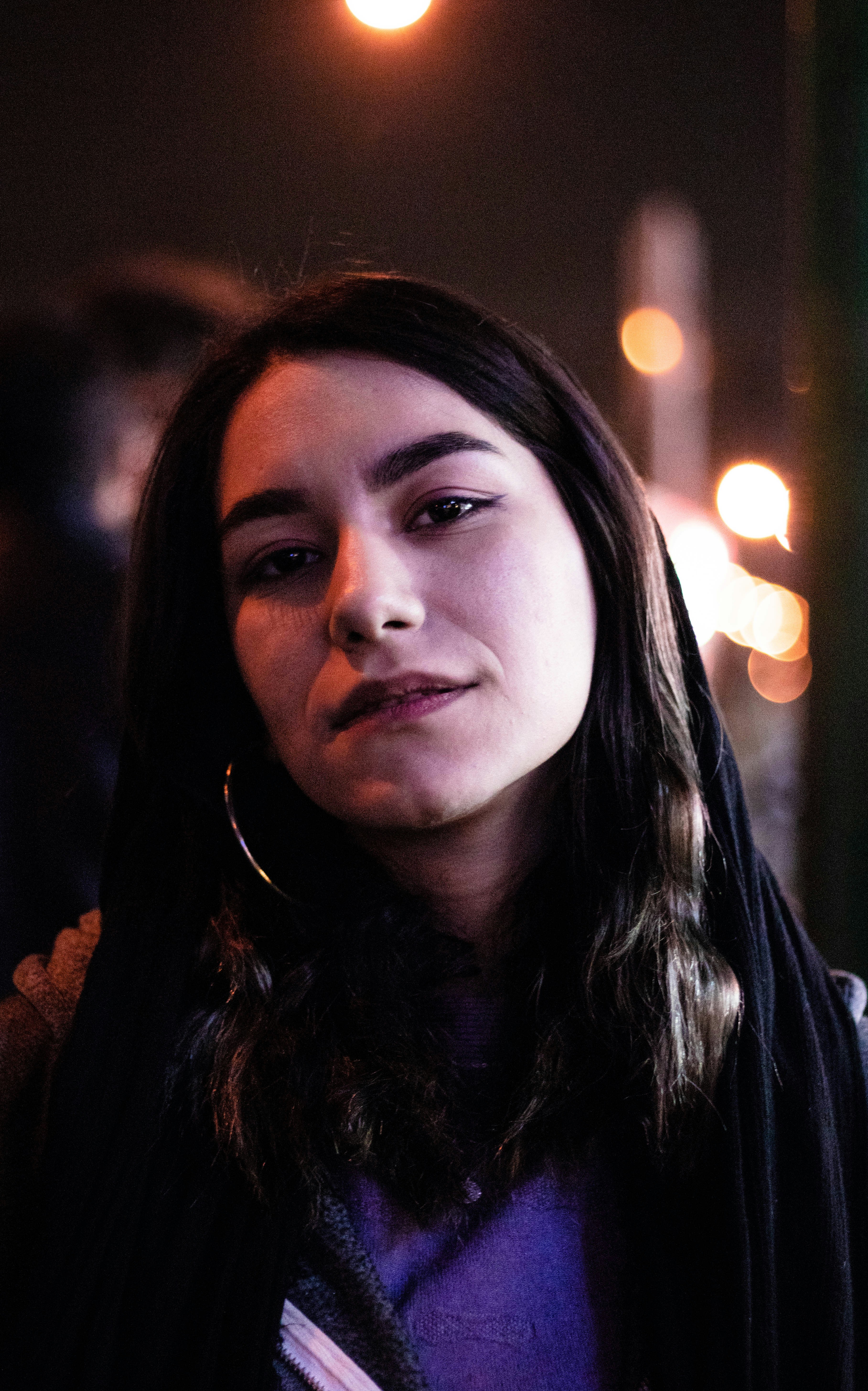 A young woman with long hair and a black scarf gazes confidently at the camera amidst a softly blurred urban backdrop. The warm glow of streetlights adds depth to the scene.