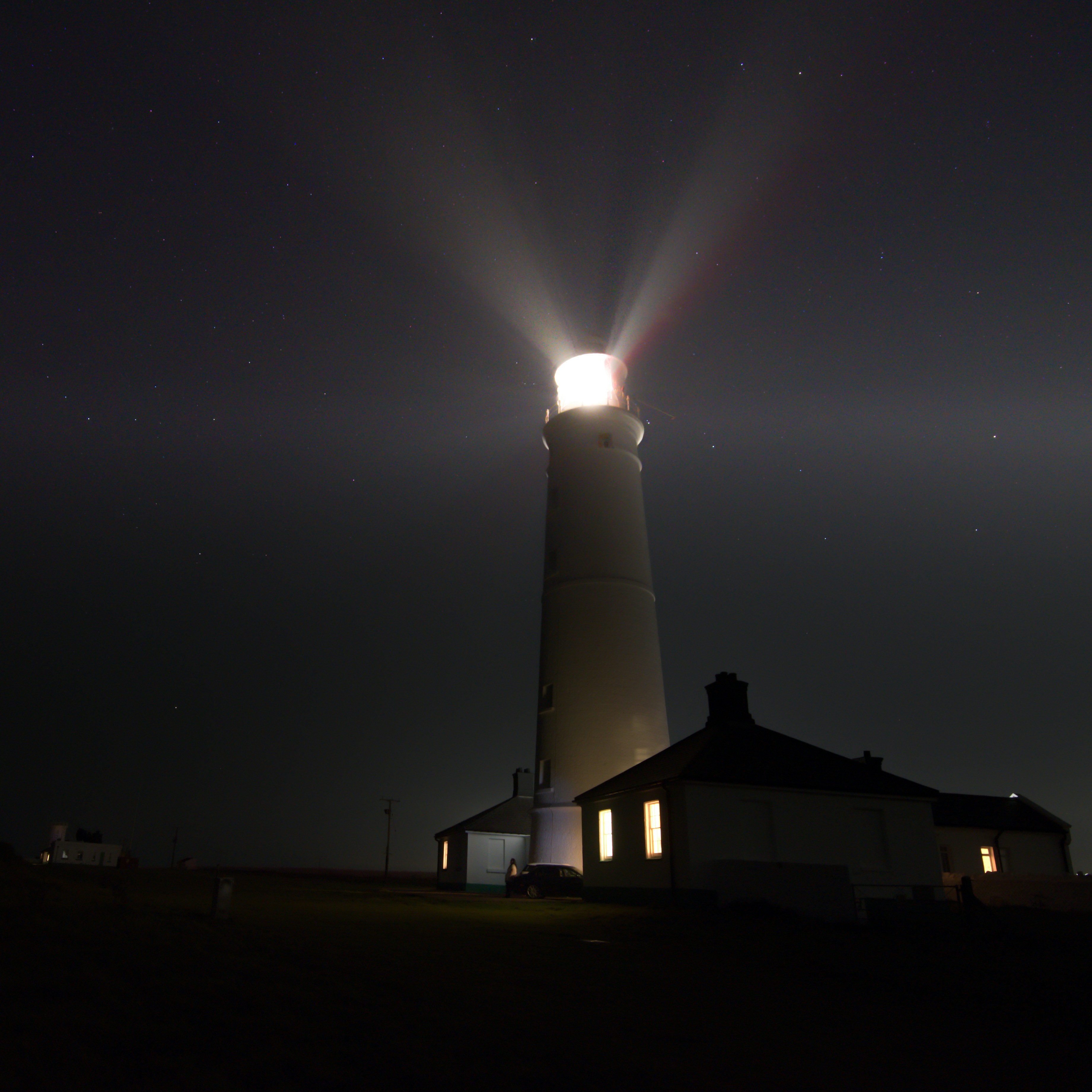 A lighthouse stands tall against a starry night, its beam cutting through the darkness. The surrounding buildings are subtly illuminated.