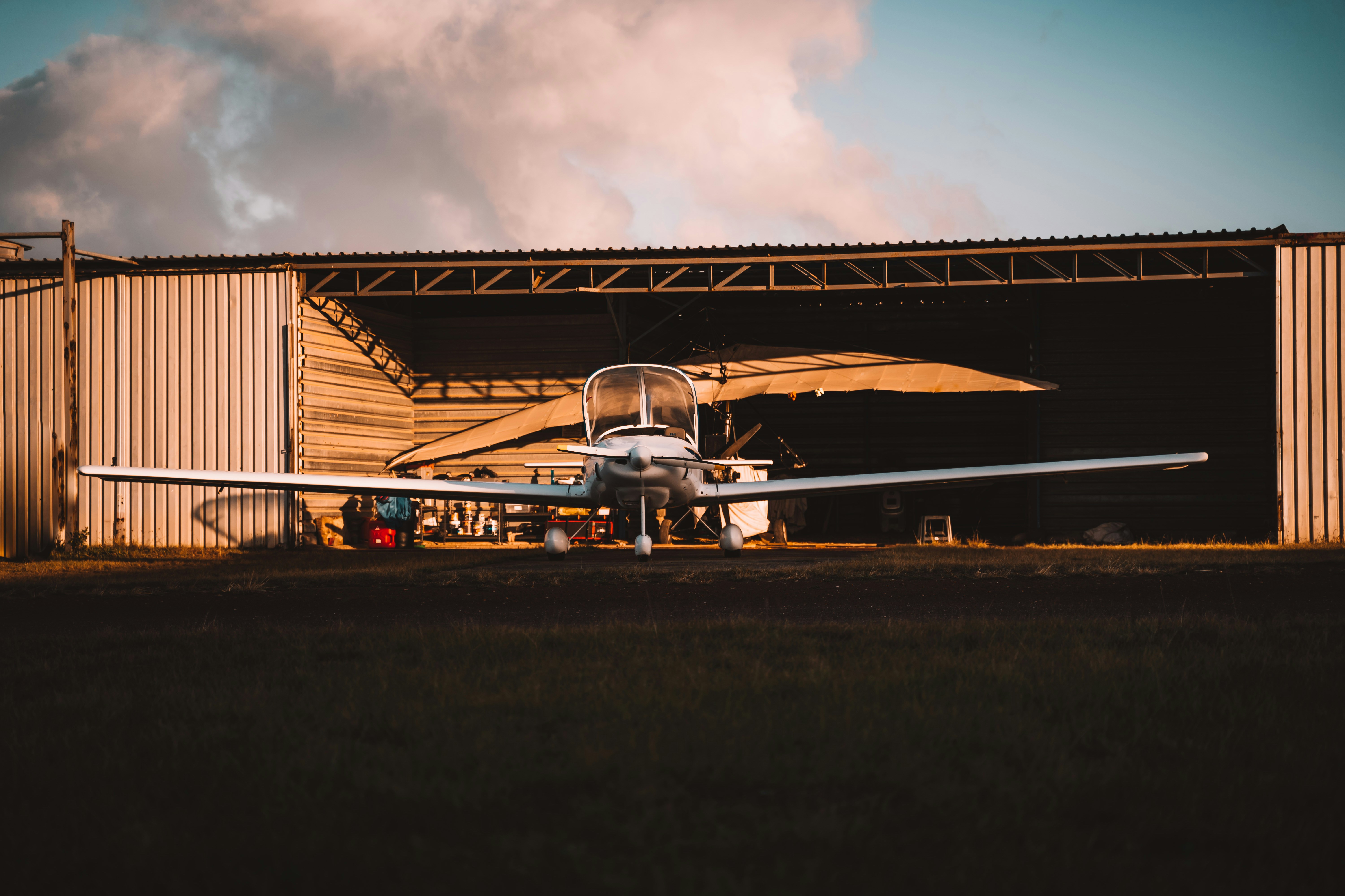 Single-engine aircraft positioned in front of an open hangar, bathed in warm evening light. Tools and equipment are visible inside the hangar.