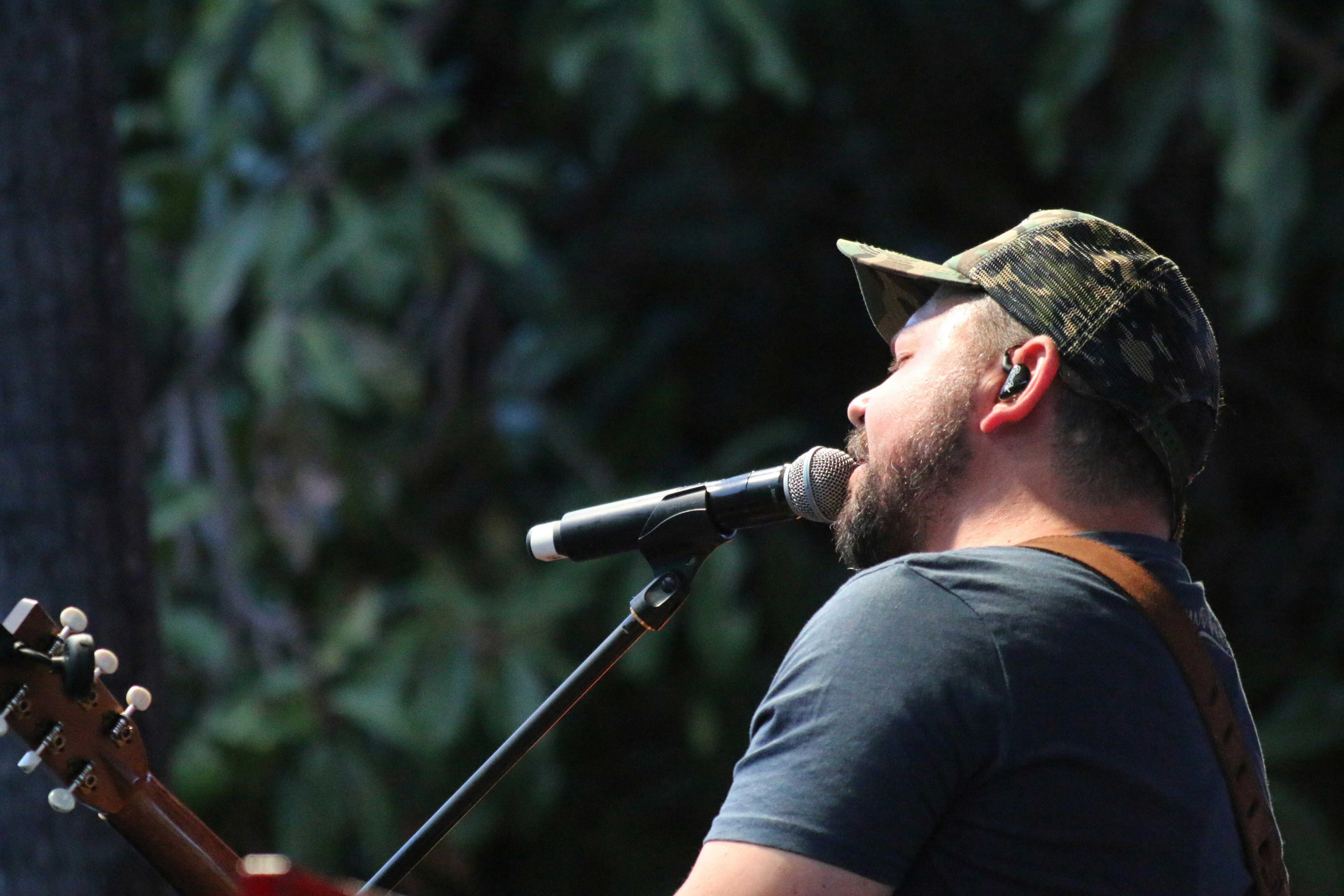 Forest Stage Serenade: A bearded guitarist-vocalist in a camo cap belts out under leafy light