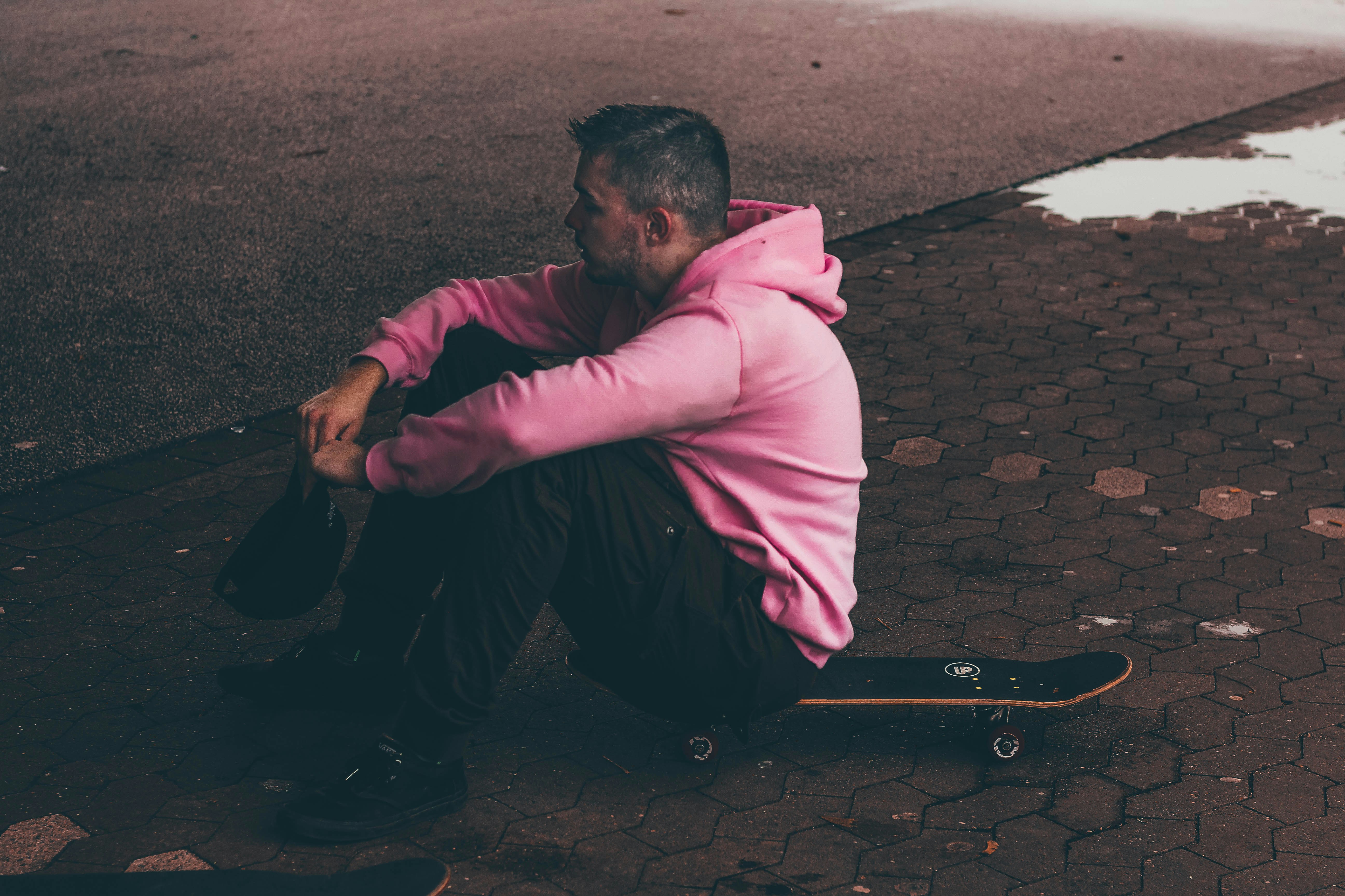 Skateboarder in a pink hoodie sitting on a skateboard, lost in thought against a textured urban backdrop.
