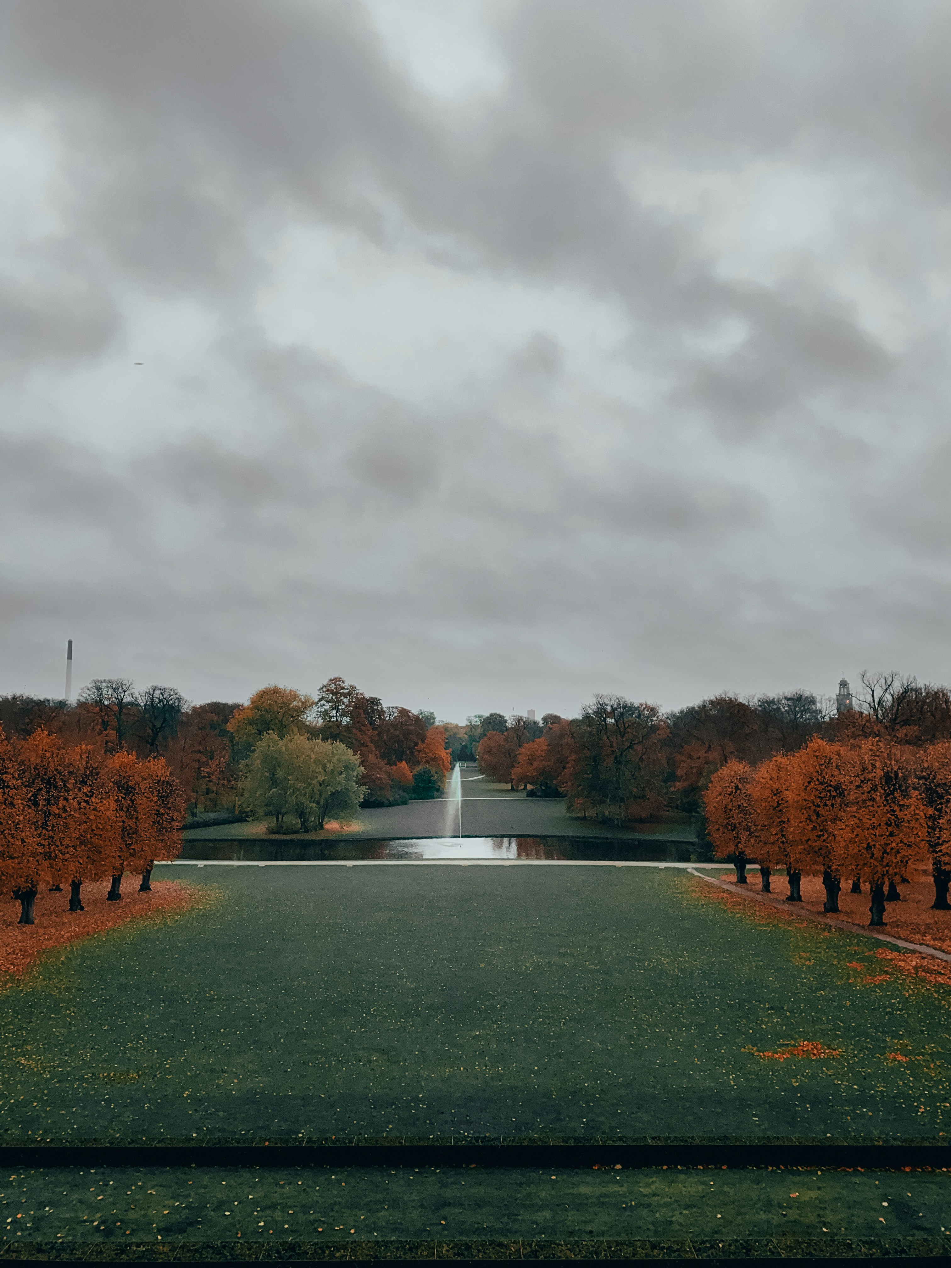Vibrant orange trees line a tranquil lake, with a fountain at the center, under a cloudy sky. The scene captures the essence of autumn's transition.