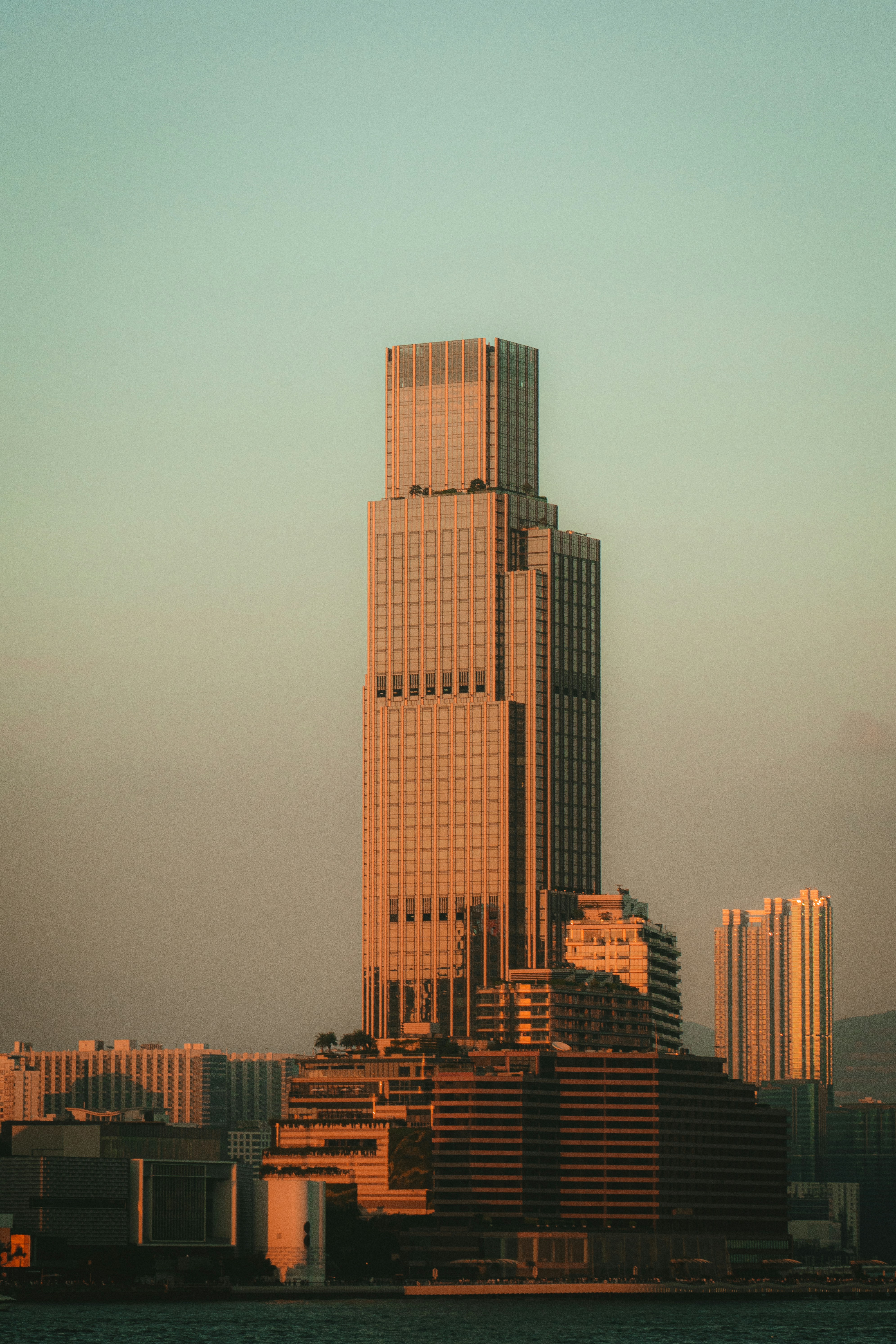 A towering skyscraper reflecting the warm hues of sunset, surrounded by modern architecture and cityscape. The structure showcases contemporary design elements.