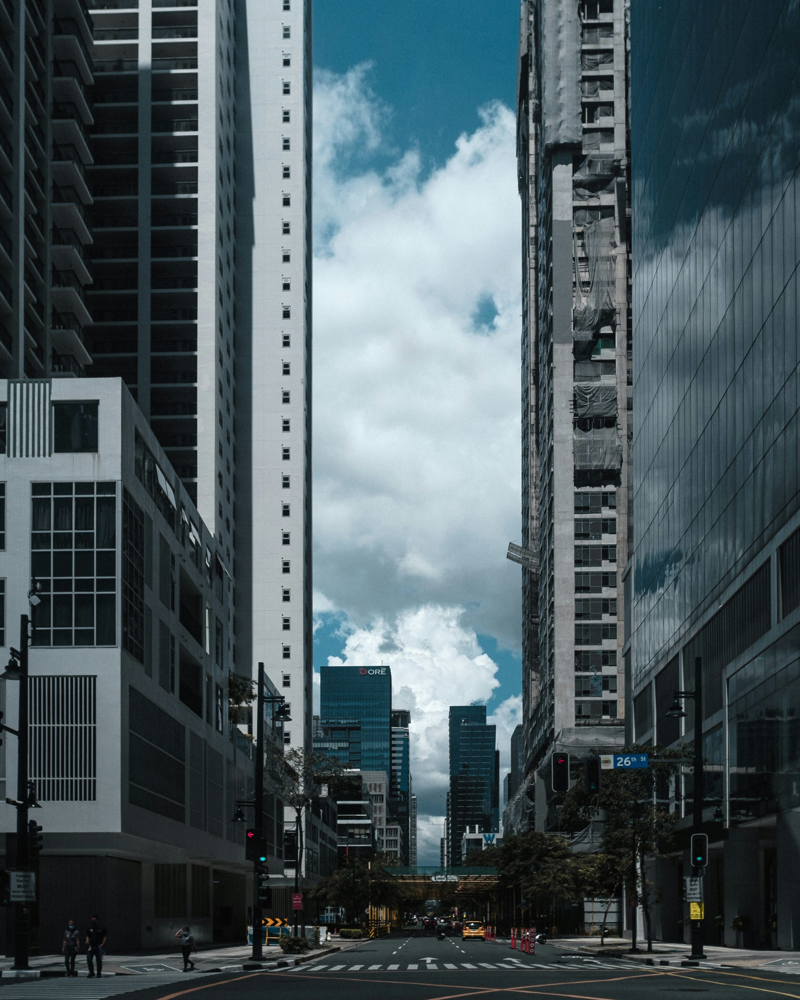 Modern skyscrapers frame a bustling city street, with clouds casting dynamic shadows overhead.