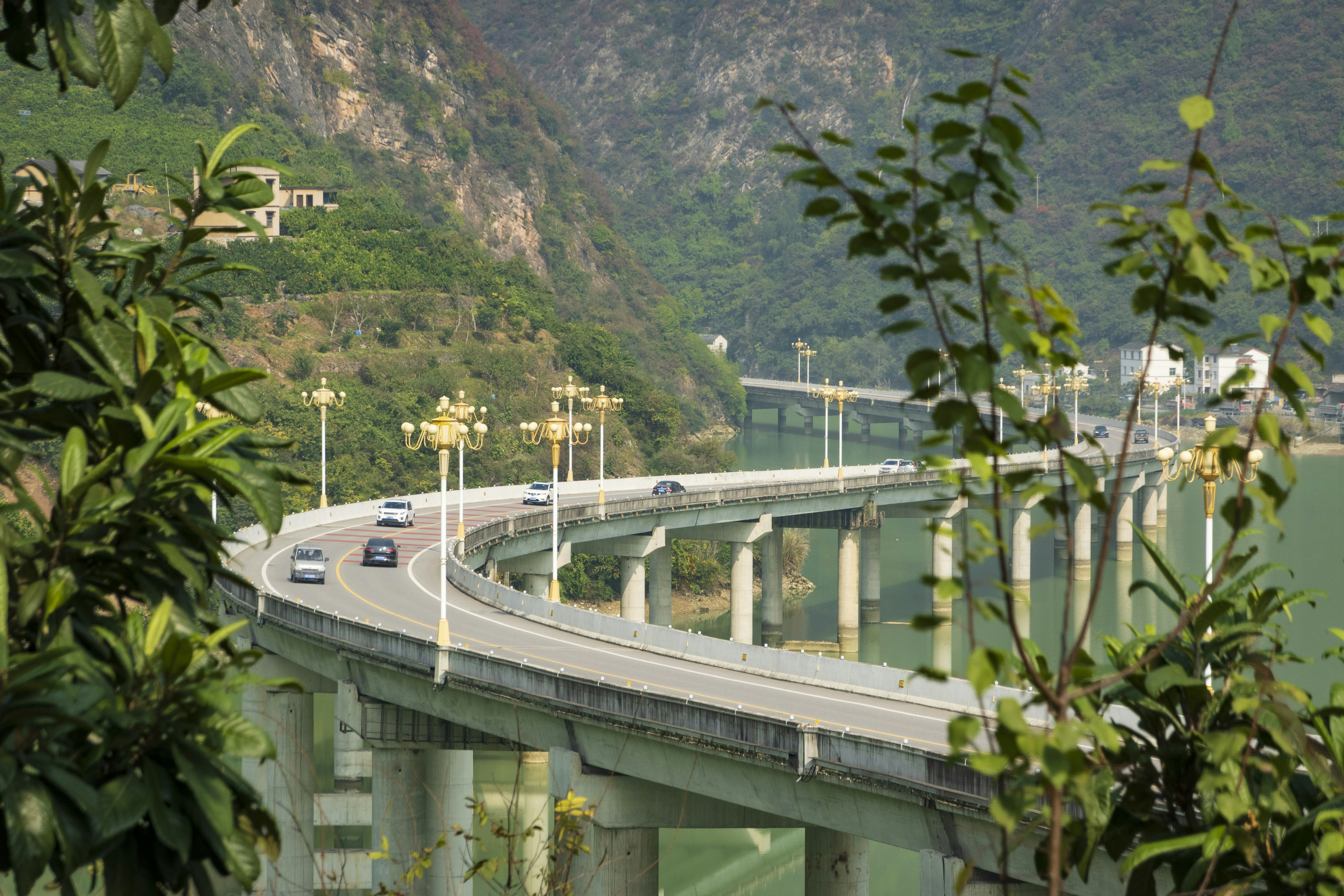 Curved highway meandering through lush greenery and mountainous terrain, with vehicles traversing the scenic route. Street lamps line the road, adding an urban touch to the natural landscape.
