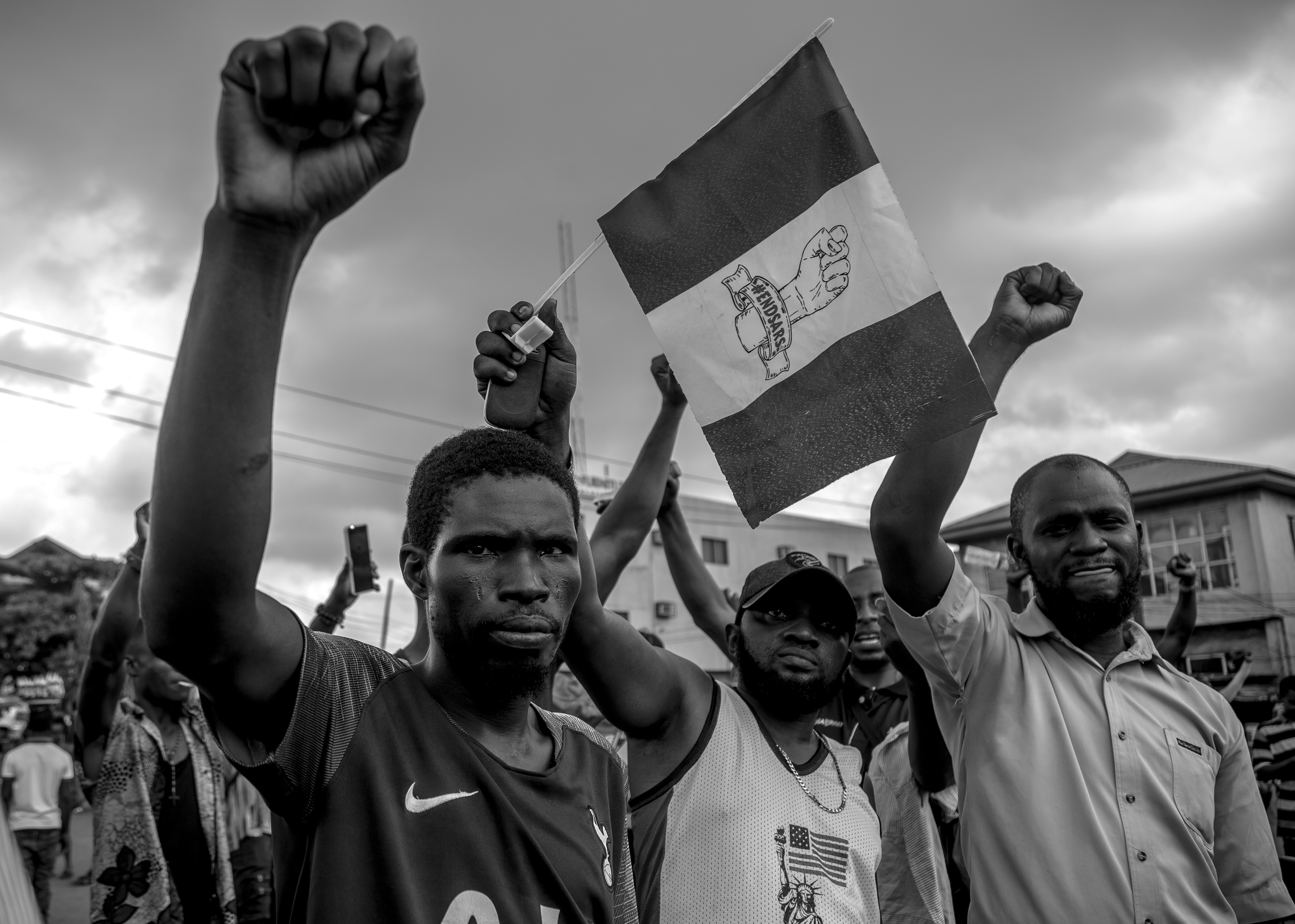 Protesters with raised fists, one holding a flag, in a grayscale cityscape.