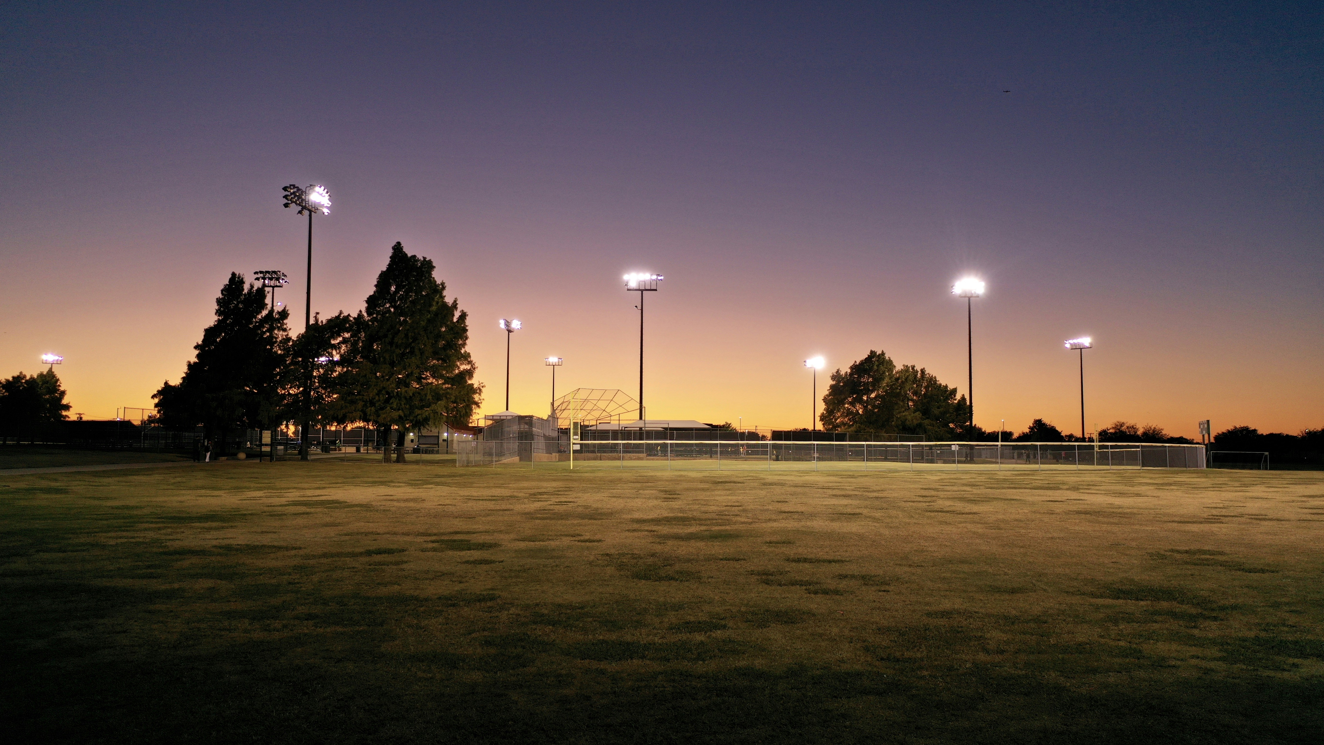 Floodlit sports field with trees silhouetted against a vibrant sunset sky.