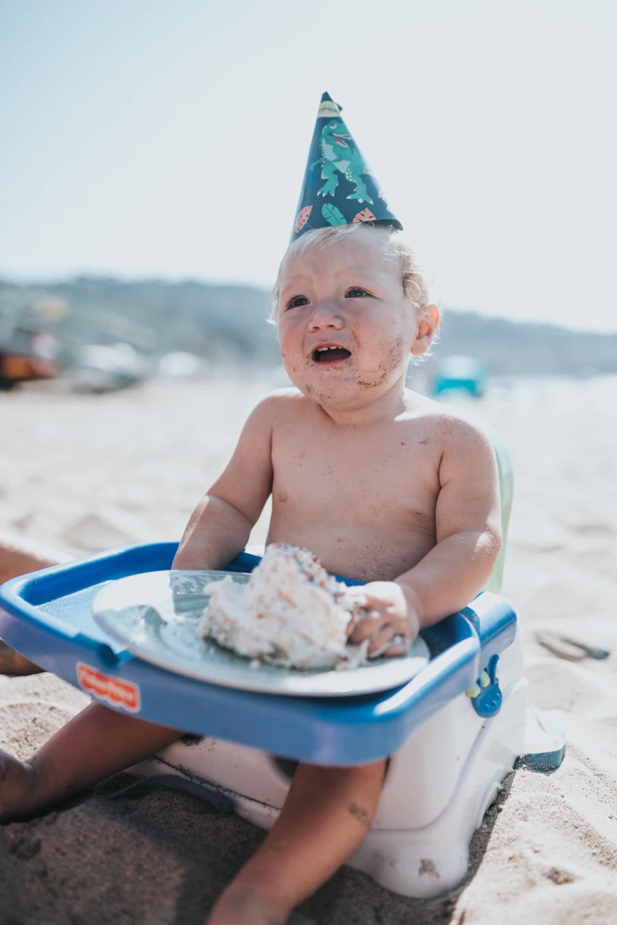 A smiling toddler sits in a blue high chair on a sandy beach, wearing a party hat and holding a piece of cake; the scene captures a celebratory moment by the shoreline. This is a candid photo featuring a child at a birthday moment.