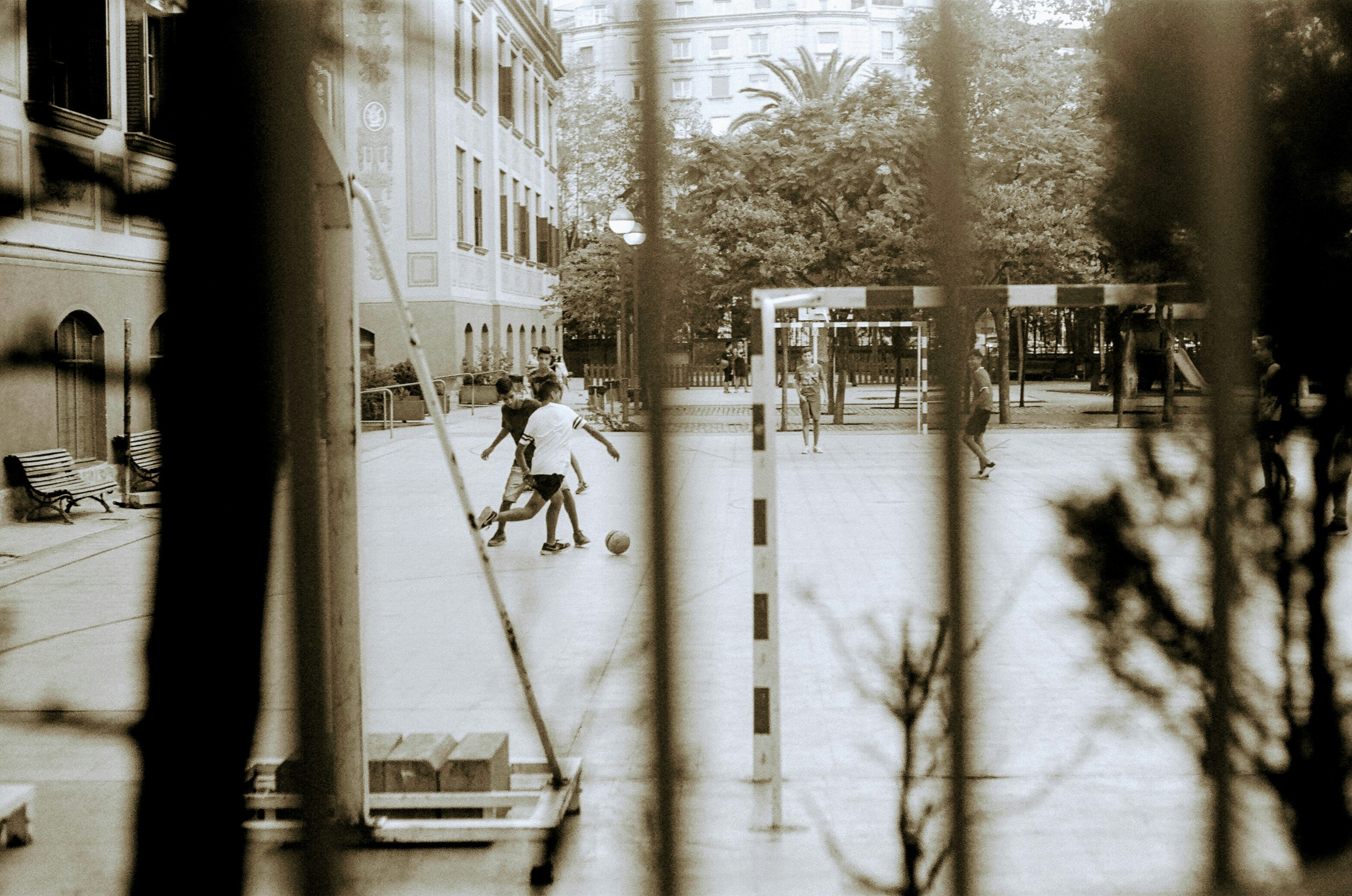 Two children engage in a spirited game of soccer on a playground, framed by the surrounding fence and greenery. The scene captures the essence of youthful playfulness.