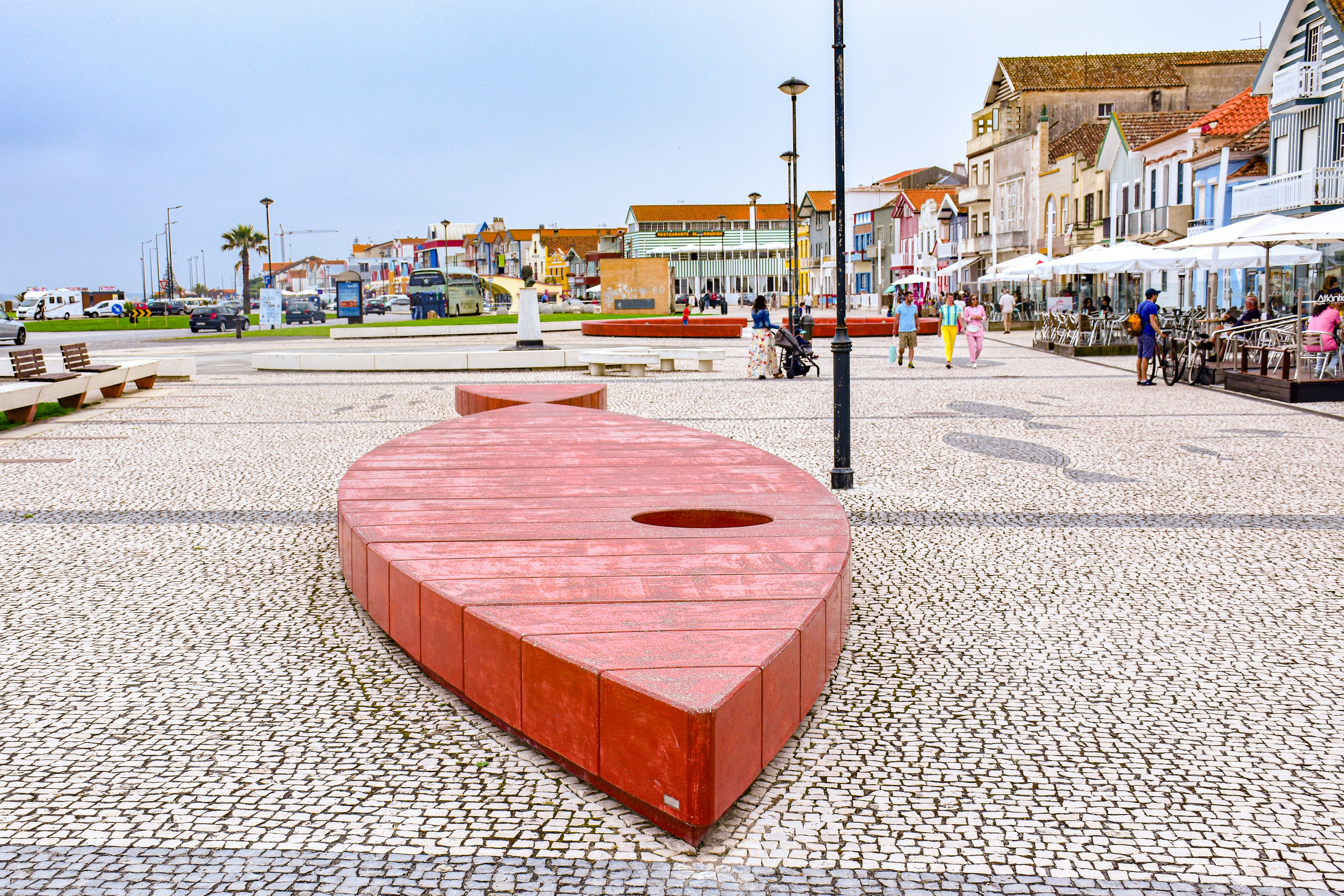 A large, red fish-shaped sculpture anchors a vibrant coastal promenade, surrounded by colorful buildings and lively outdoor cafés.